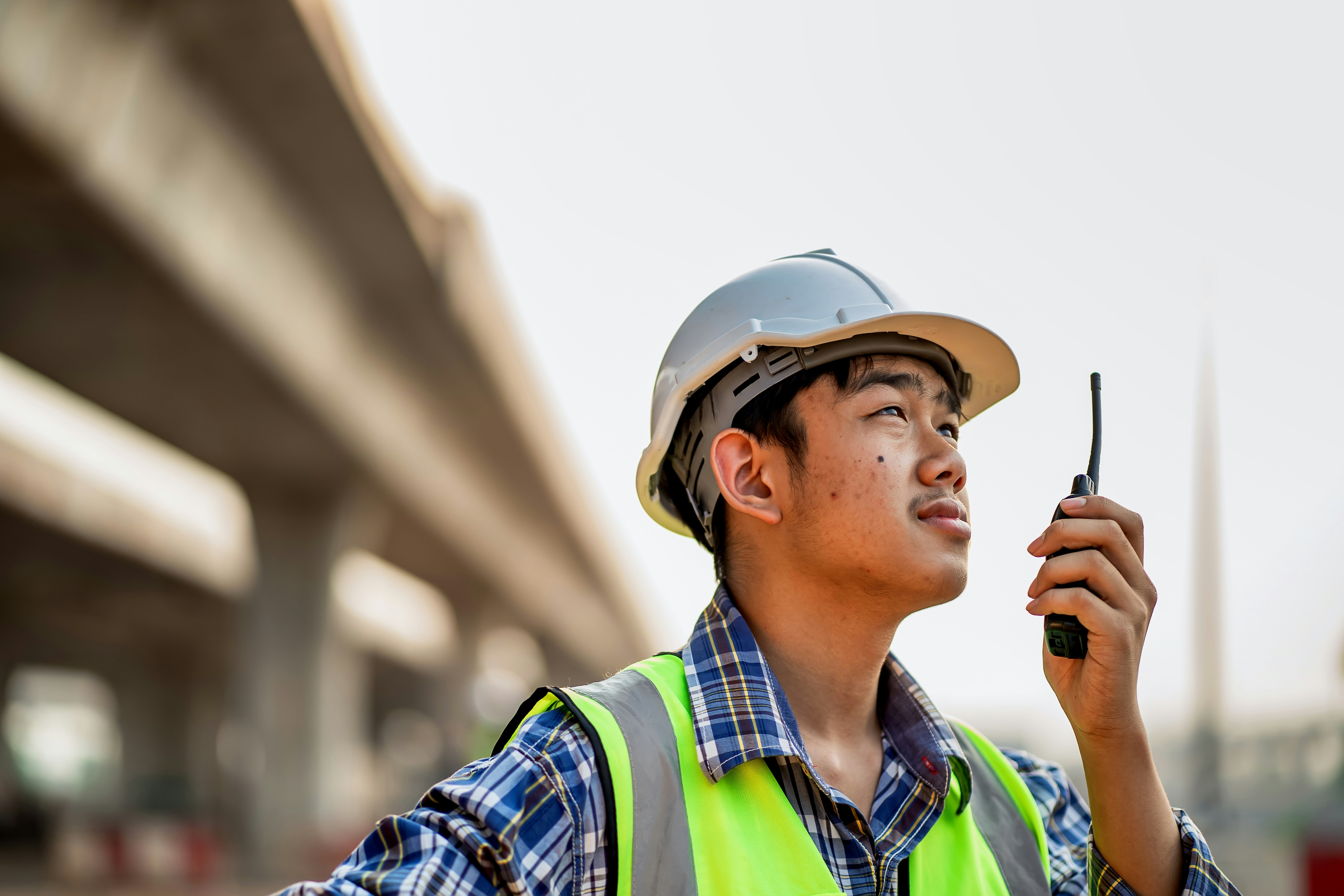 Engineer in a hard hat and high-visibility vest communicates via radio under a highway bridge.