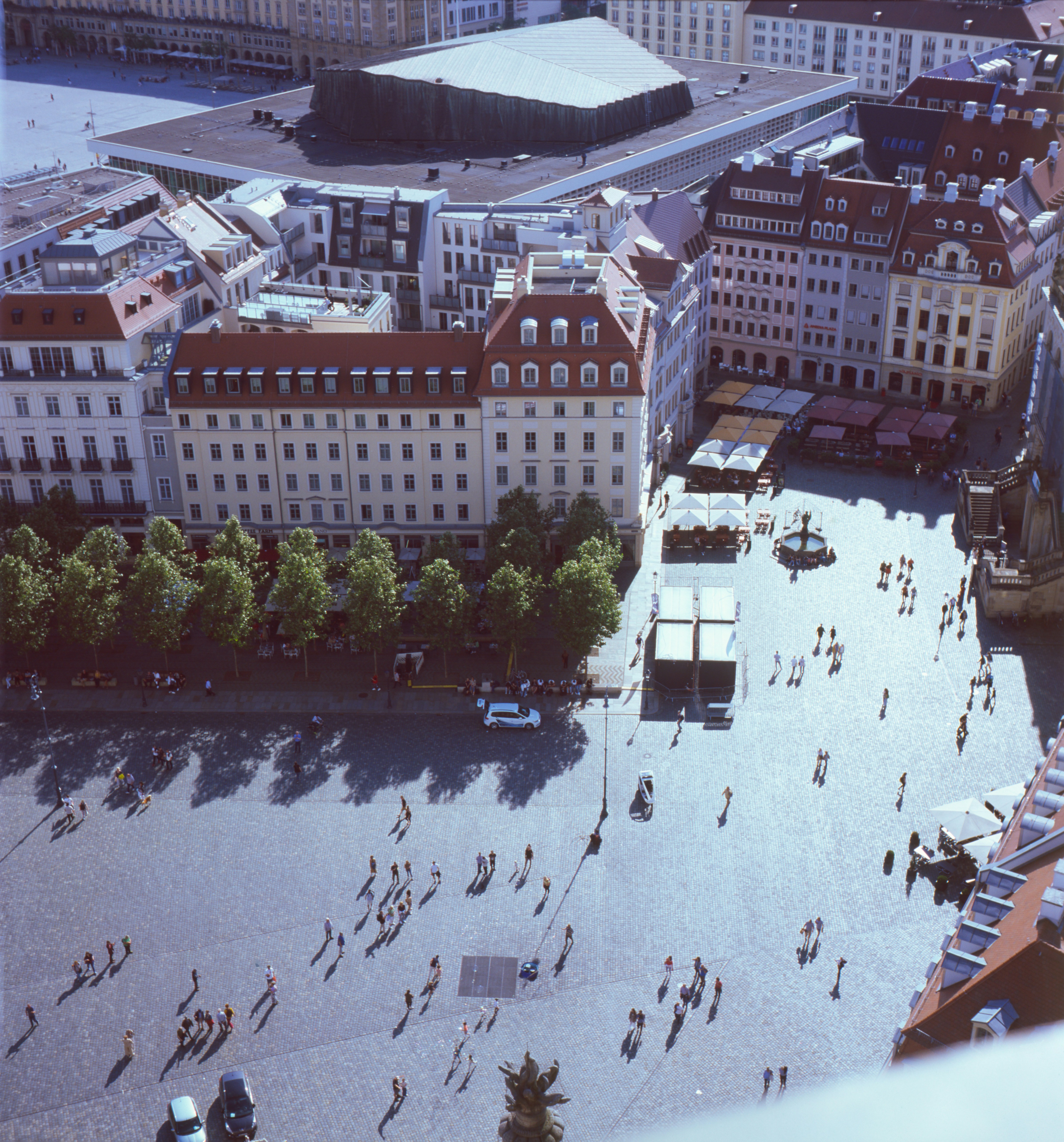 Aerial view of a bustling plaza, with people moving amidst historic architecture and lush trees. The interplay of shadows creates a dynamic atmosphere.
