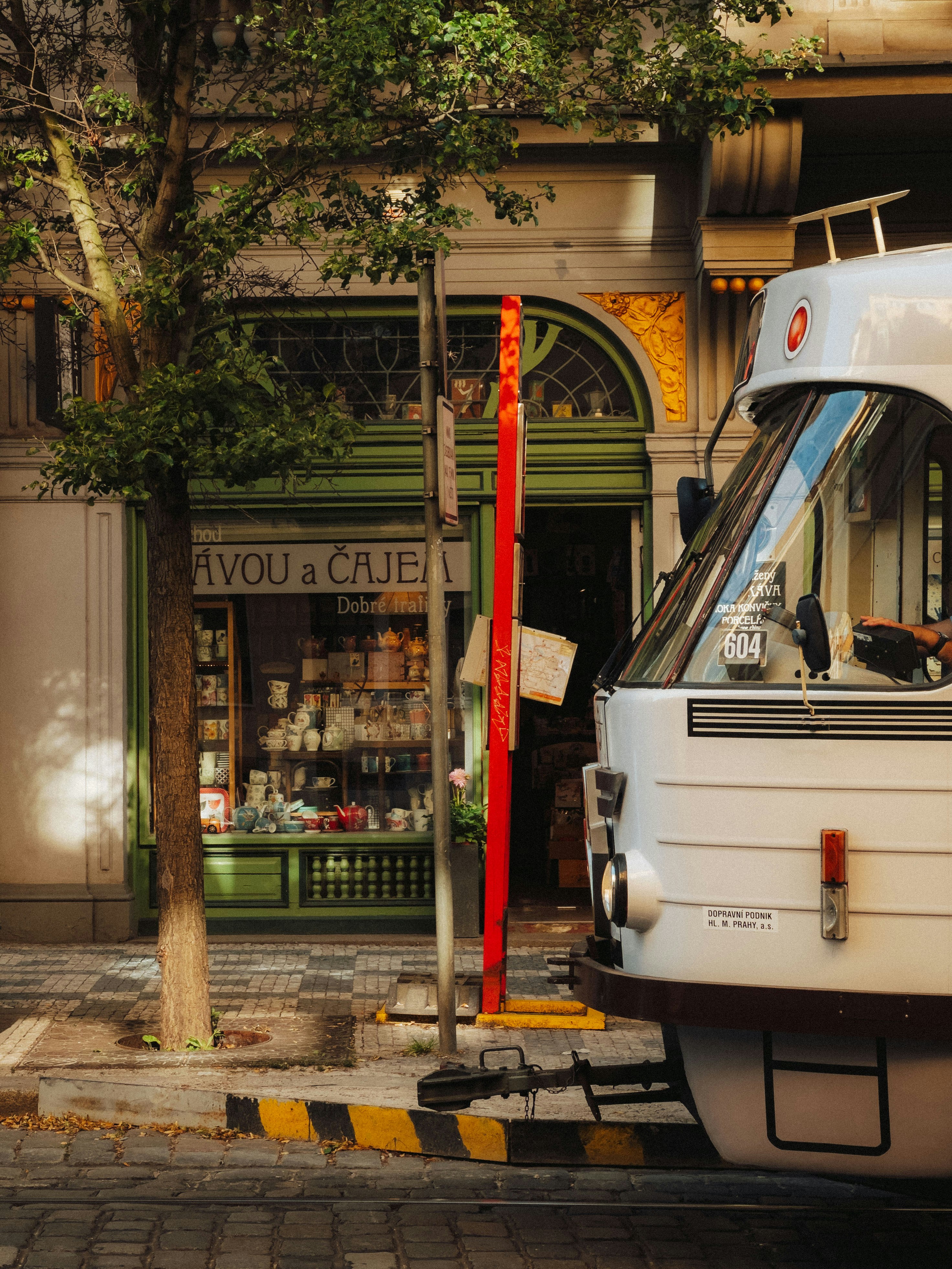 A vintage tram pauses beside a charming storefront adorned with colorful wares. Lush greenery frames the scene, blending urban life with nature.