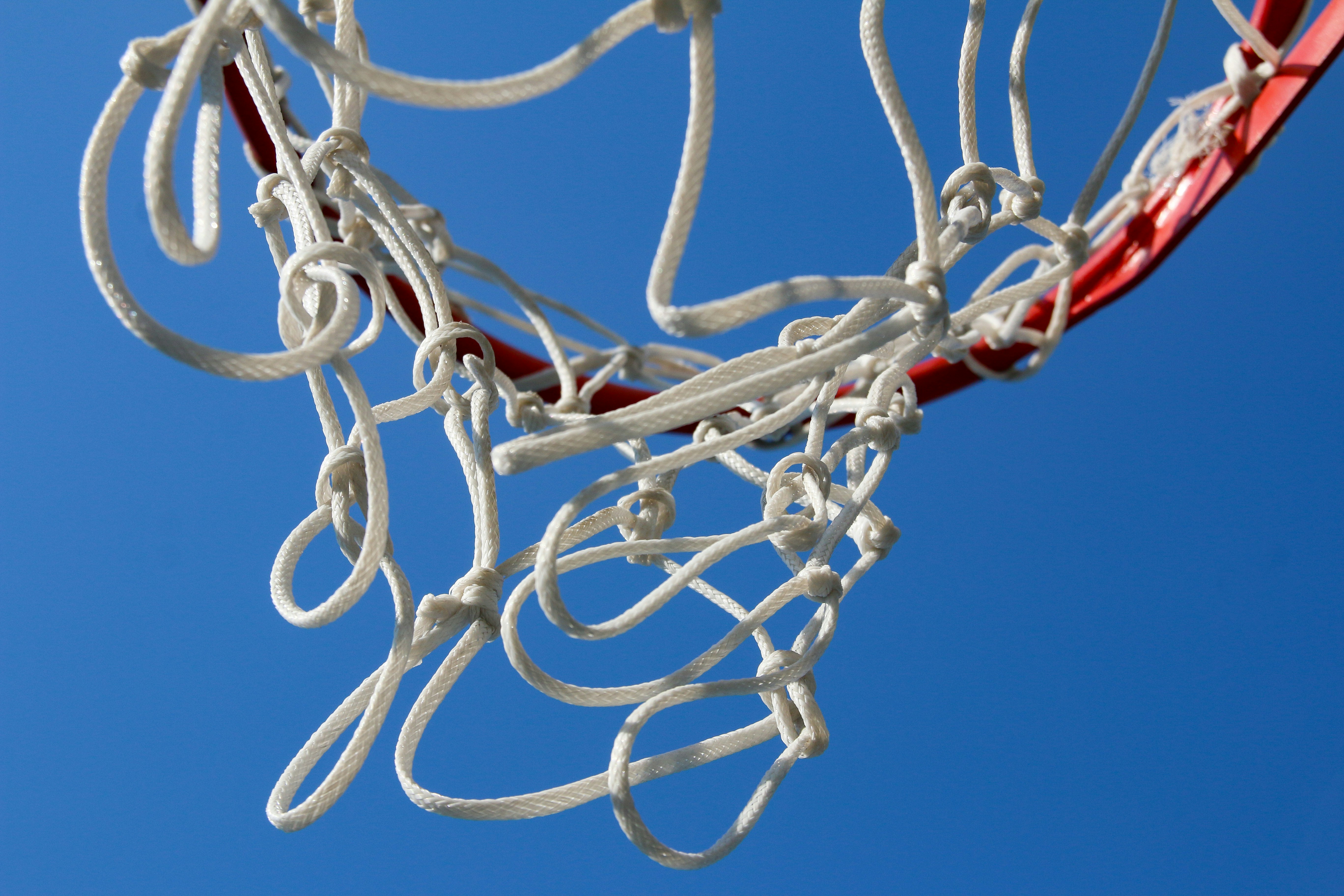 Close-up of basketball net tangled against a bright blue sky, capturing the texture of the ropes and the red rim threads. The composition emphasizes the net’s loops and negative space.