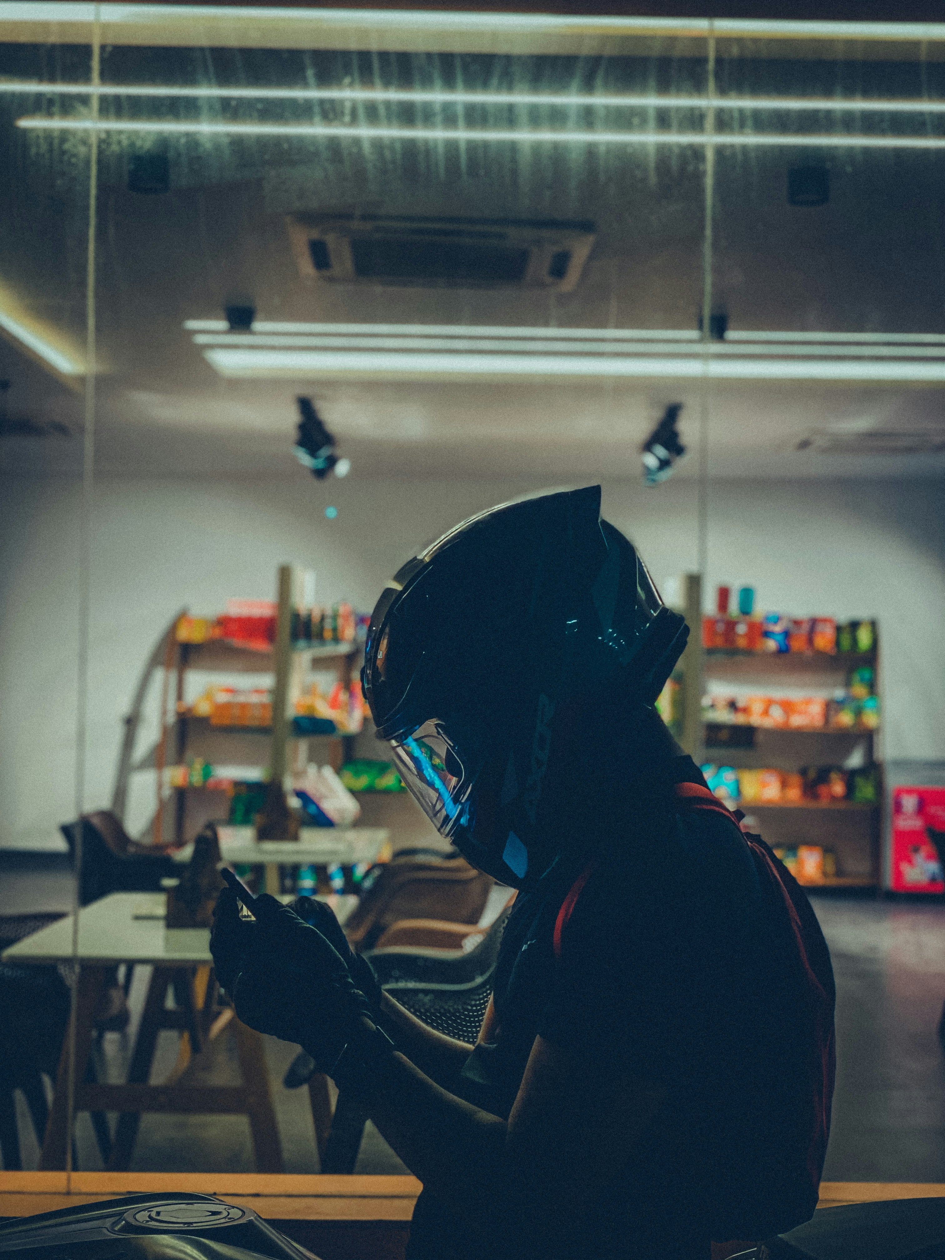 Helmeted silhouette in a glass-front shop, lit by the snack-shelf glow