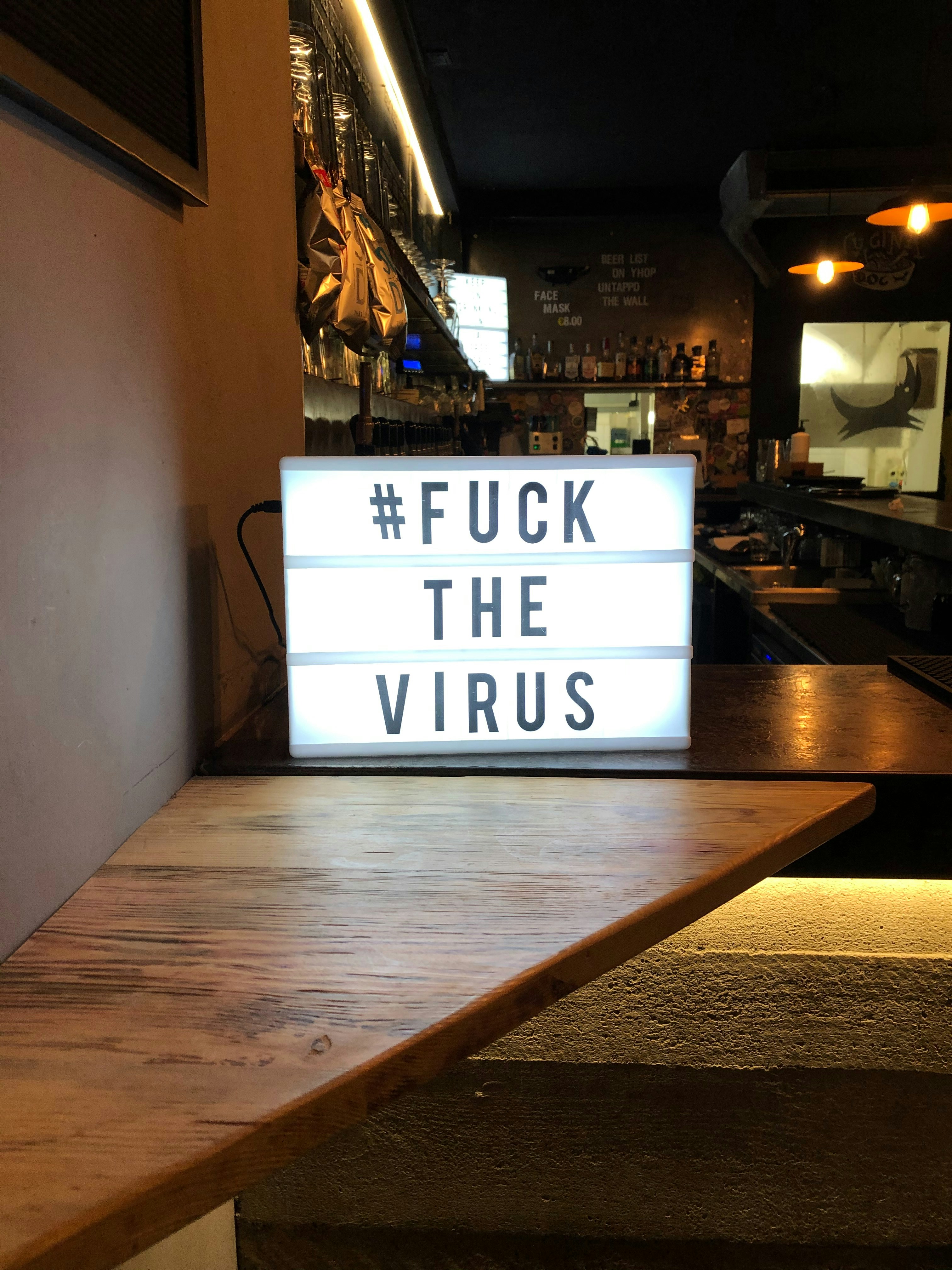 Illuminated sign reading '#FUCK THE VIRUS' placed on a wooden bar top in a dimly lit setting.
