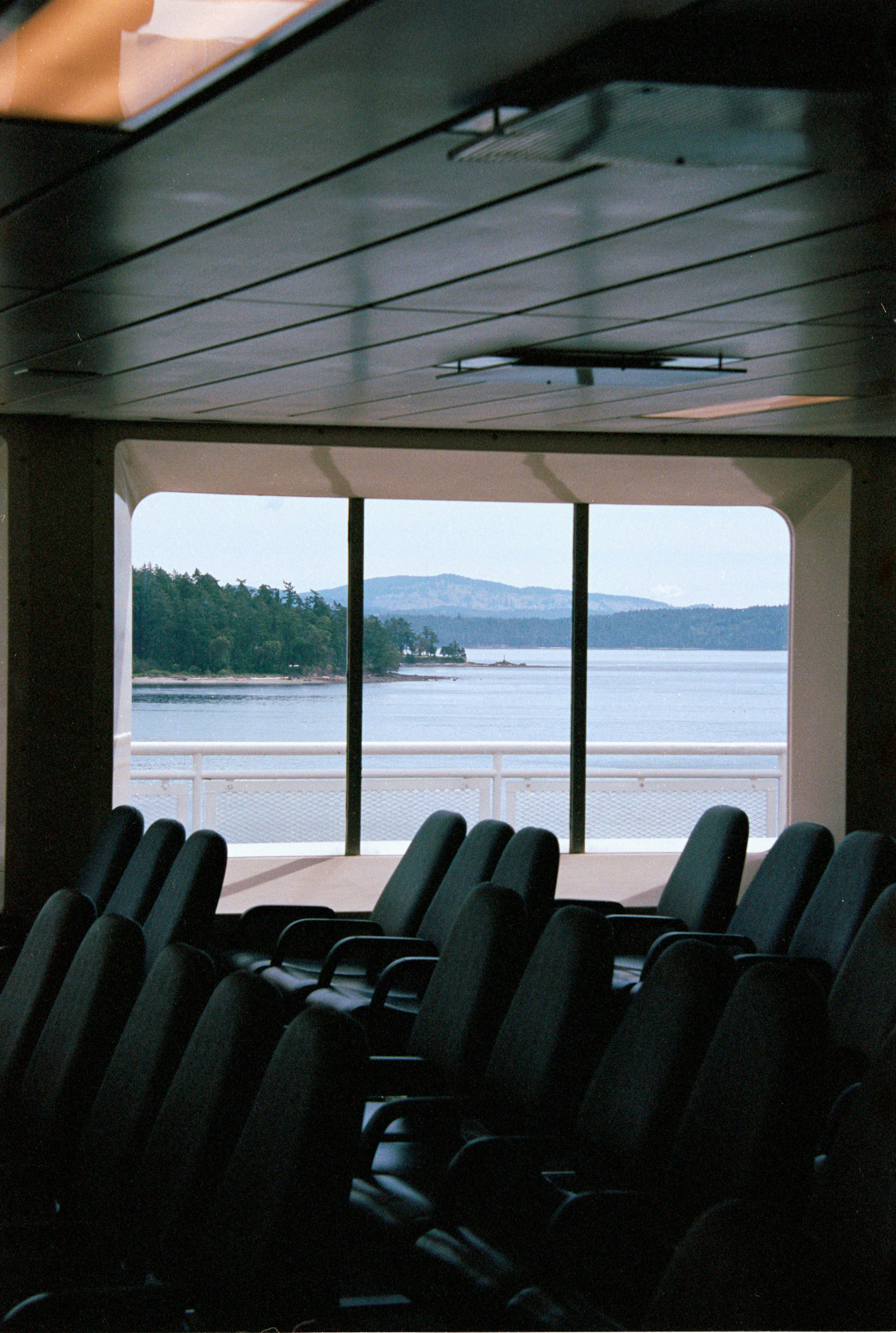 Rows of empty seats inside a ferry with large windows revealing a tranquil seascape and distant hills.
