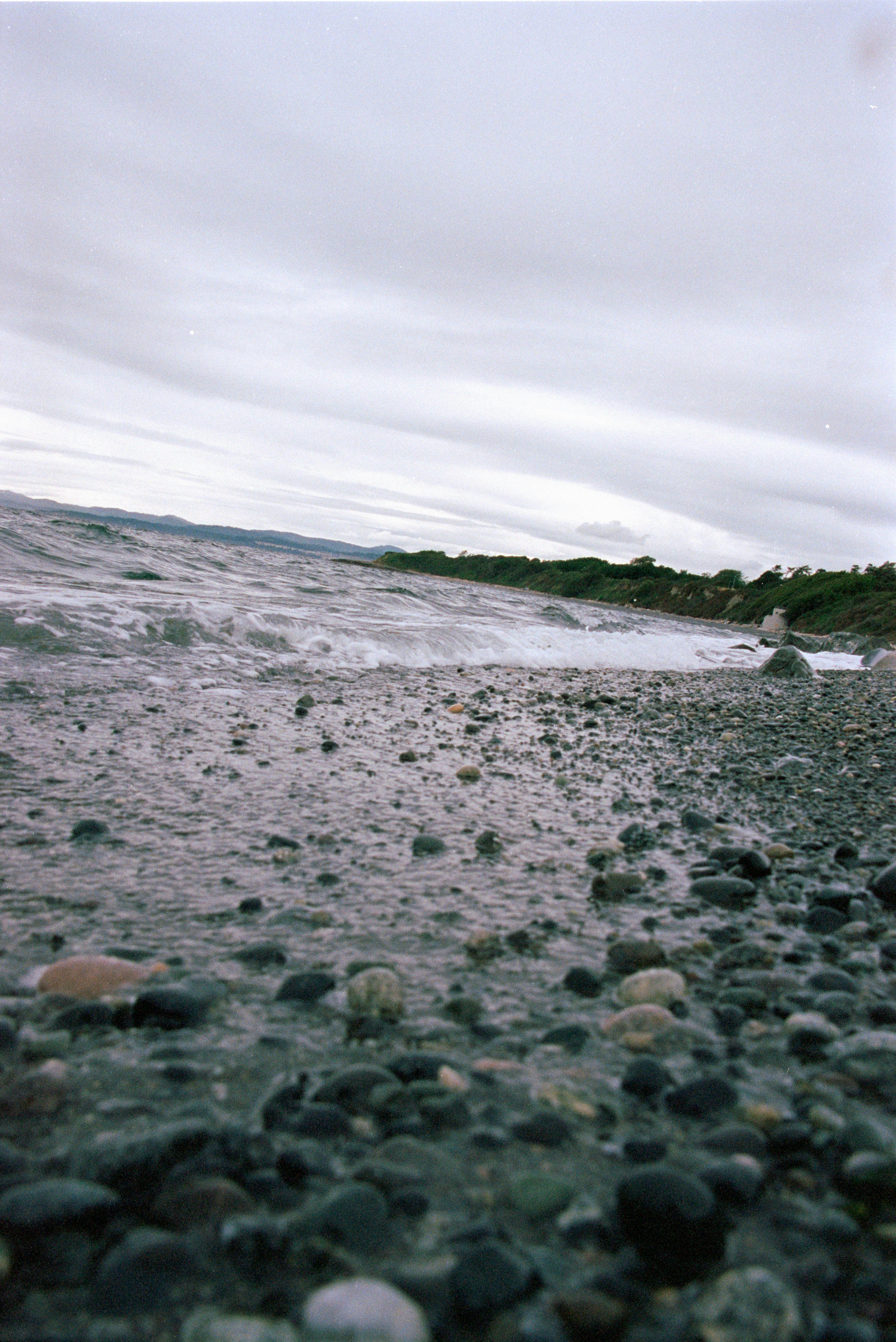 Pebble beach photograph with smooth stones in the foreground and gentle waves rolling onto shore under an overcast sky. The composition emphasizes texture of pebbles and the tranquil coastal scene.