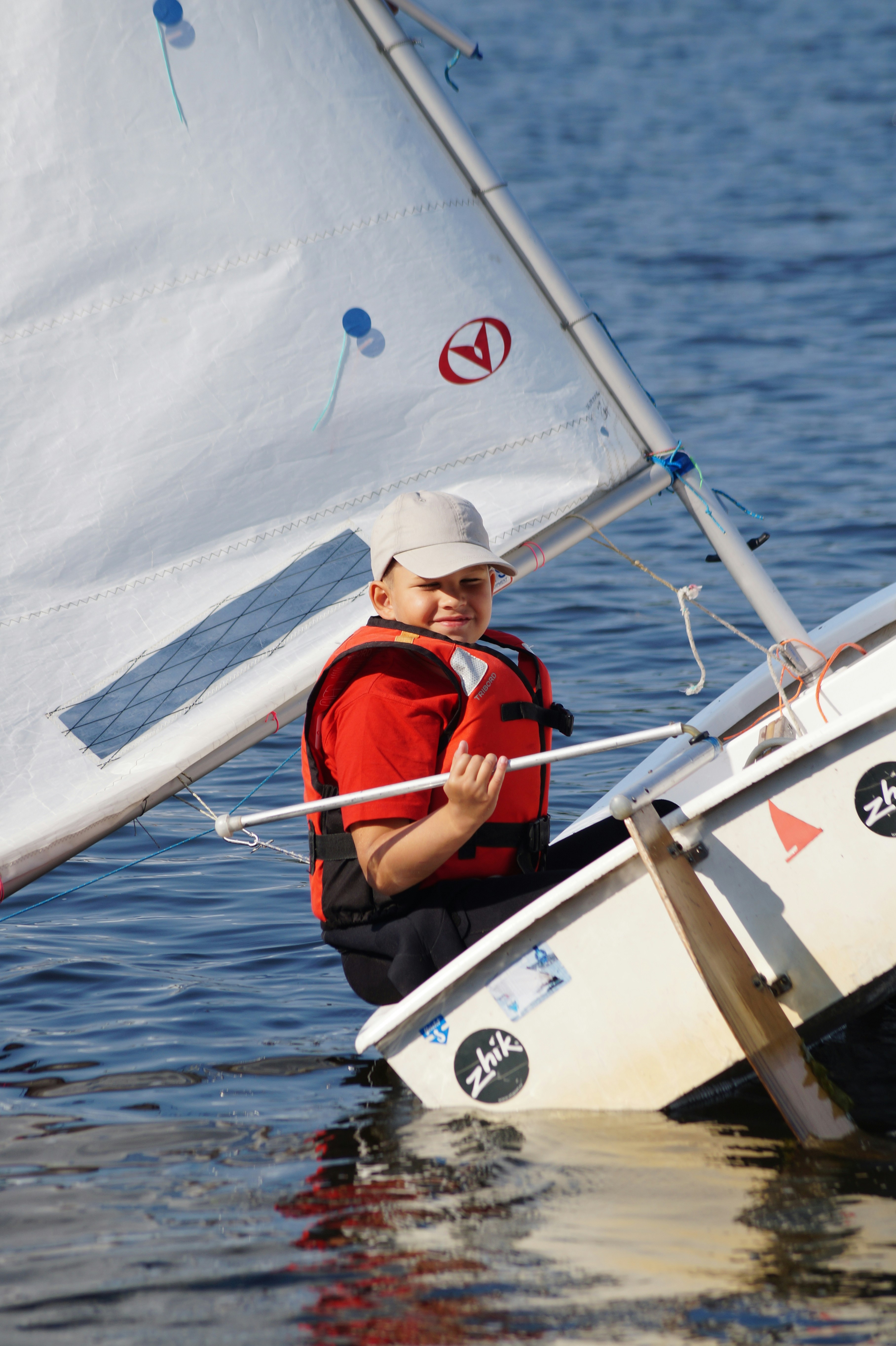 Lil Helmsman in a Red Life Jacket Navigates a White-Sailed Dinghy on Quiet Blue Waters