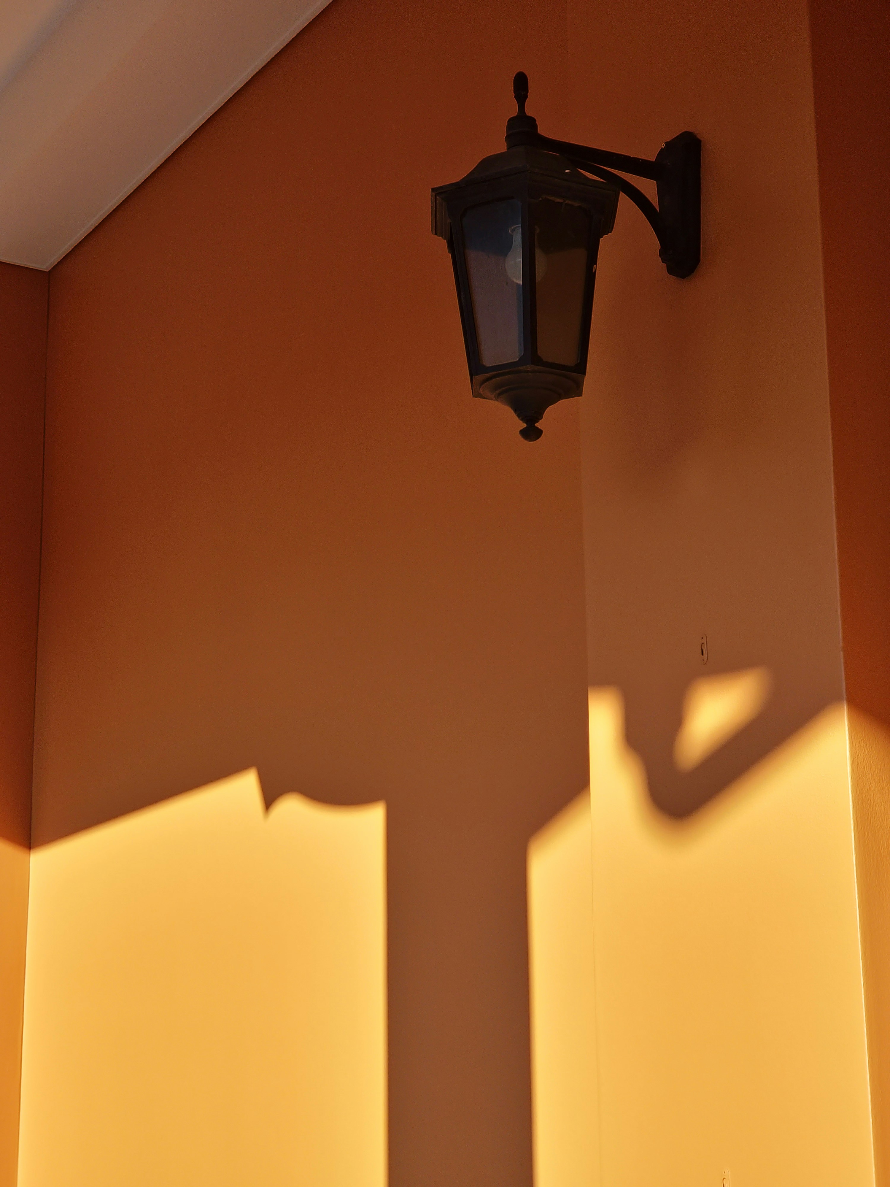 Photograph of a warm orange exterior wall with a hanging lantern, casting bold shadows. It emphasizes light geometry and wall texture.