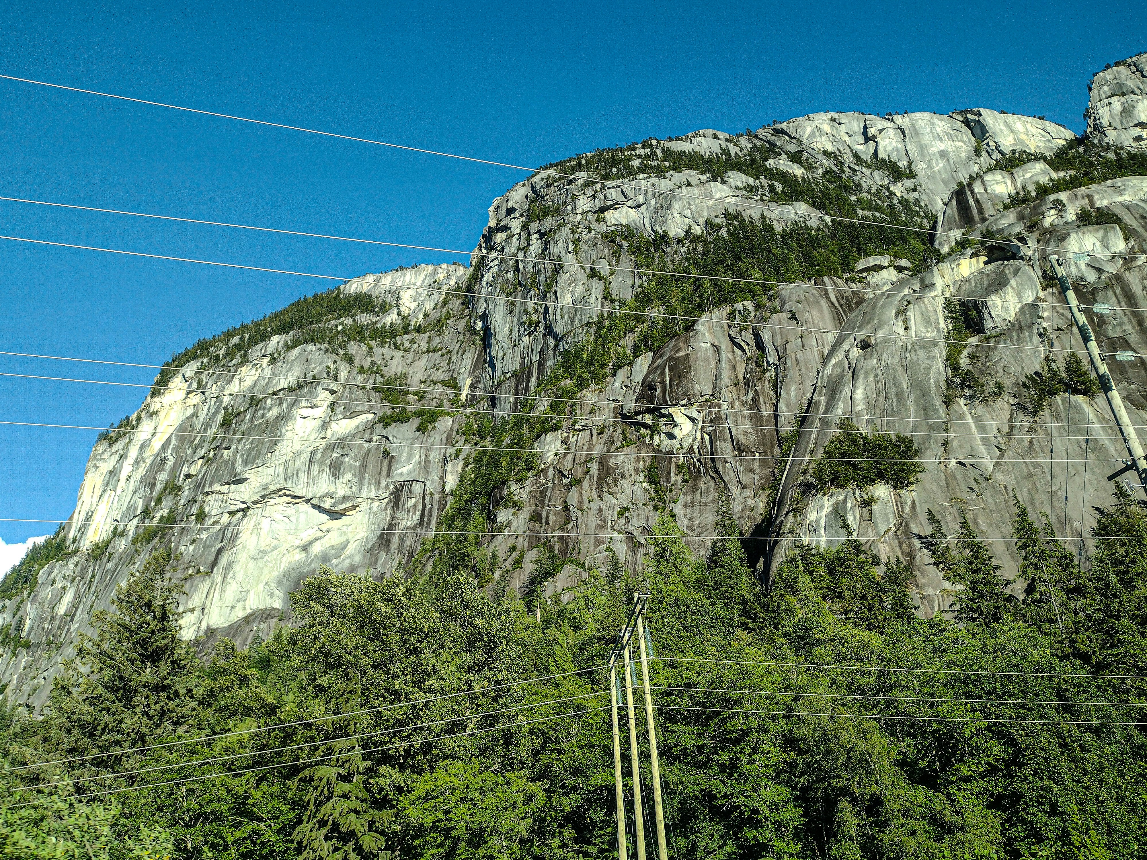 Rocky cliff face with overhead power lines and a forested base under a bright blue sky; landscape photograph.