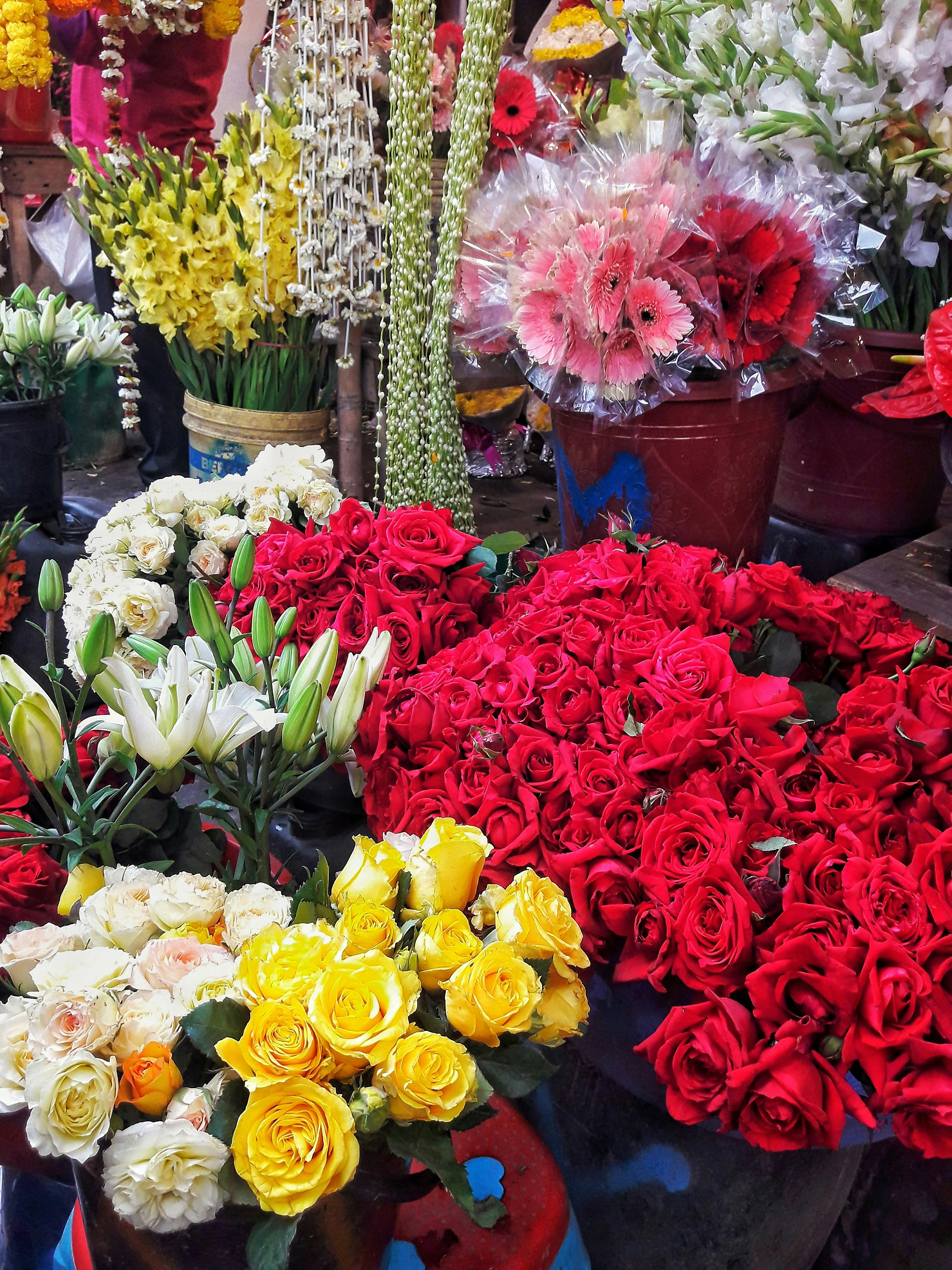 Vibrant red, yellow, and white roses fill a crowded market stall, accented by lilies and greenery in baskets and pots. The composition captures a lively floral display at a street market.
