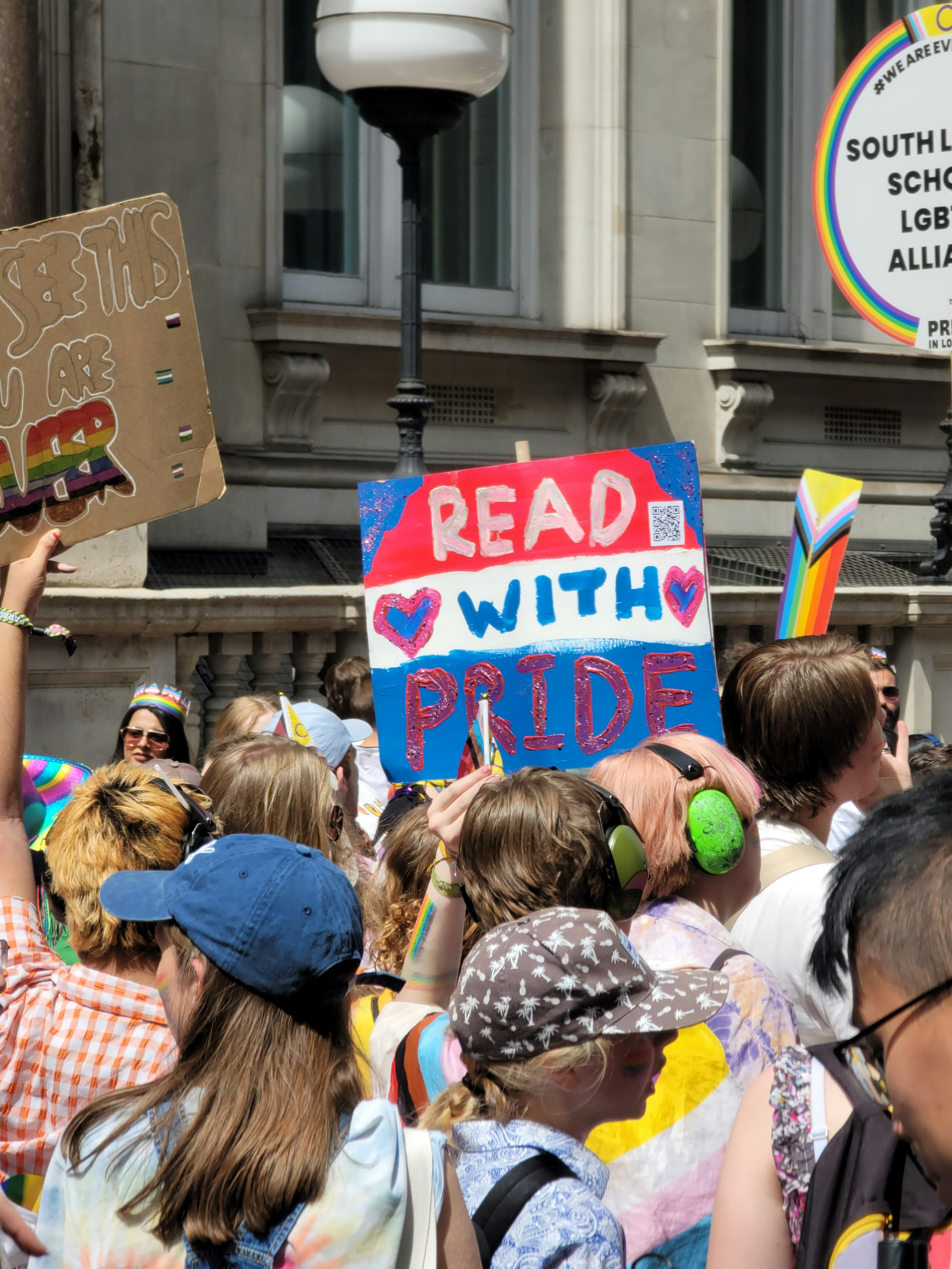 Crowd at a pride parade holding colorful signs, including one that reads 'Read with Pride.'