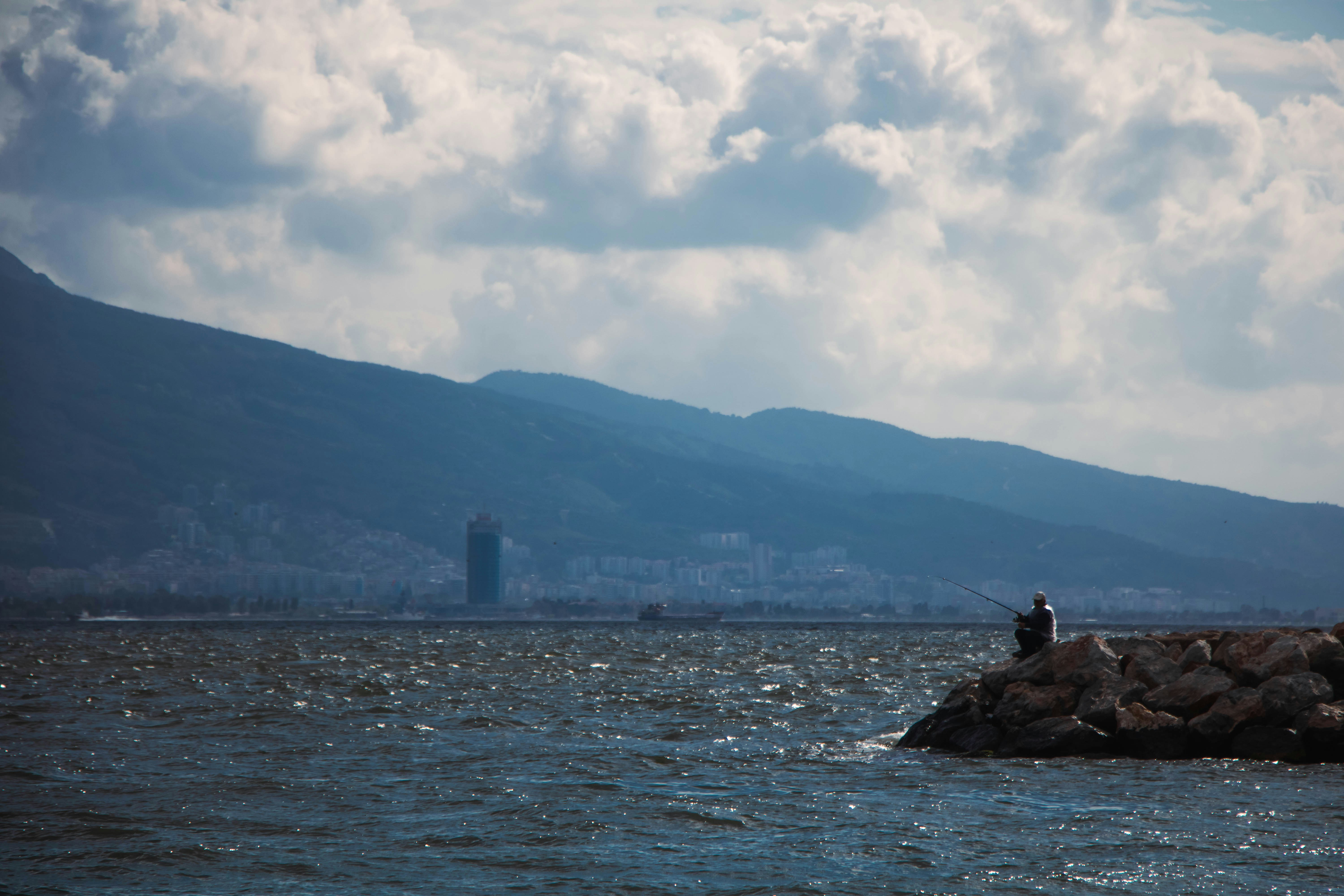 Jetty Sentinel: A Solitary Figure Framed by Sea, Mountains, and Distant City