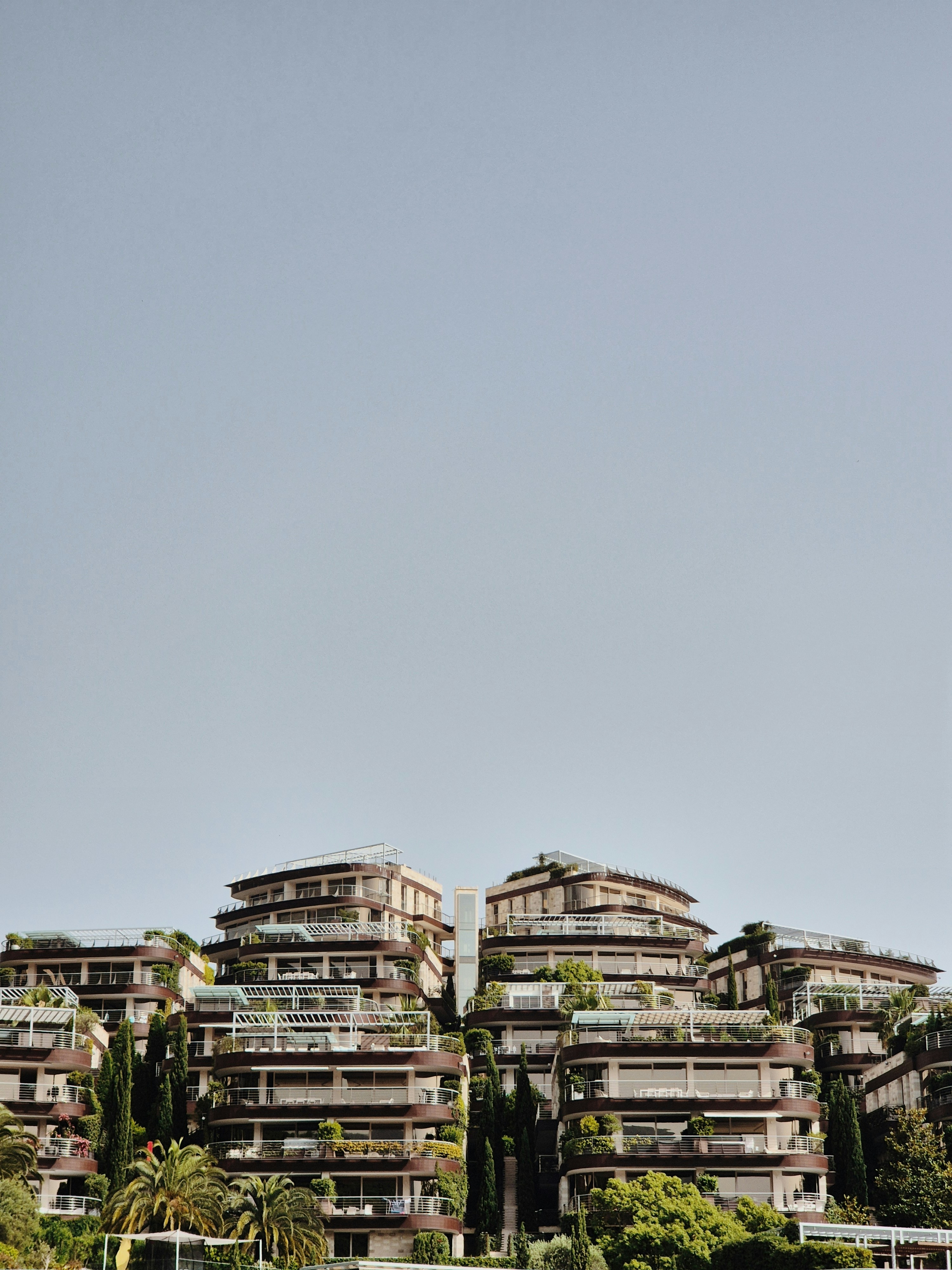 A photograph of tiered hillside apartment buildings with abundant balconies and greenery rising beneath a clear blue sky. The composition foregrounds the architectural cluster against the expansive sky.