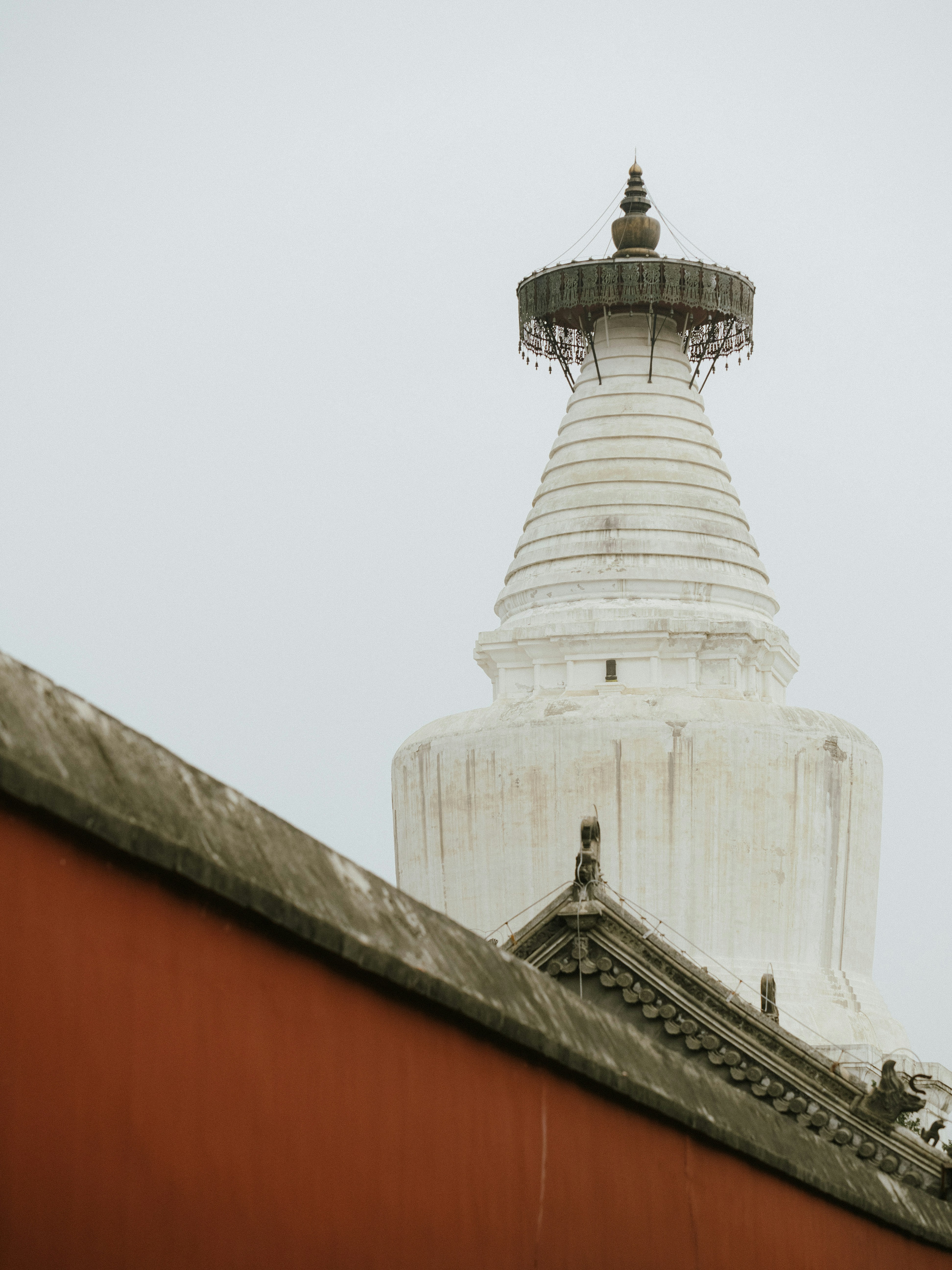 A white stupa with tiered rings rises over a red temple wall, its ornate crown detailing visible against a pale sky. The composition emphasizes architectural contrast and sacred geometry.