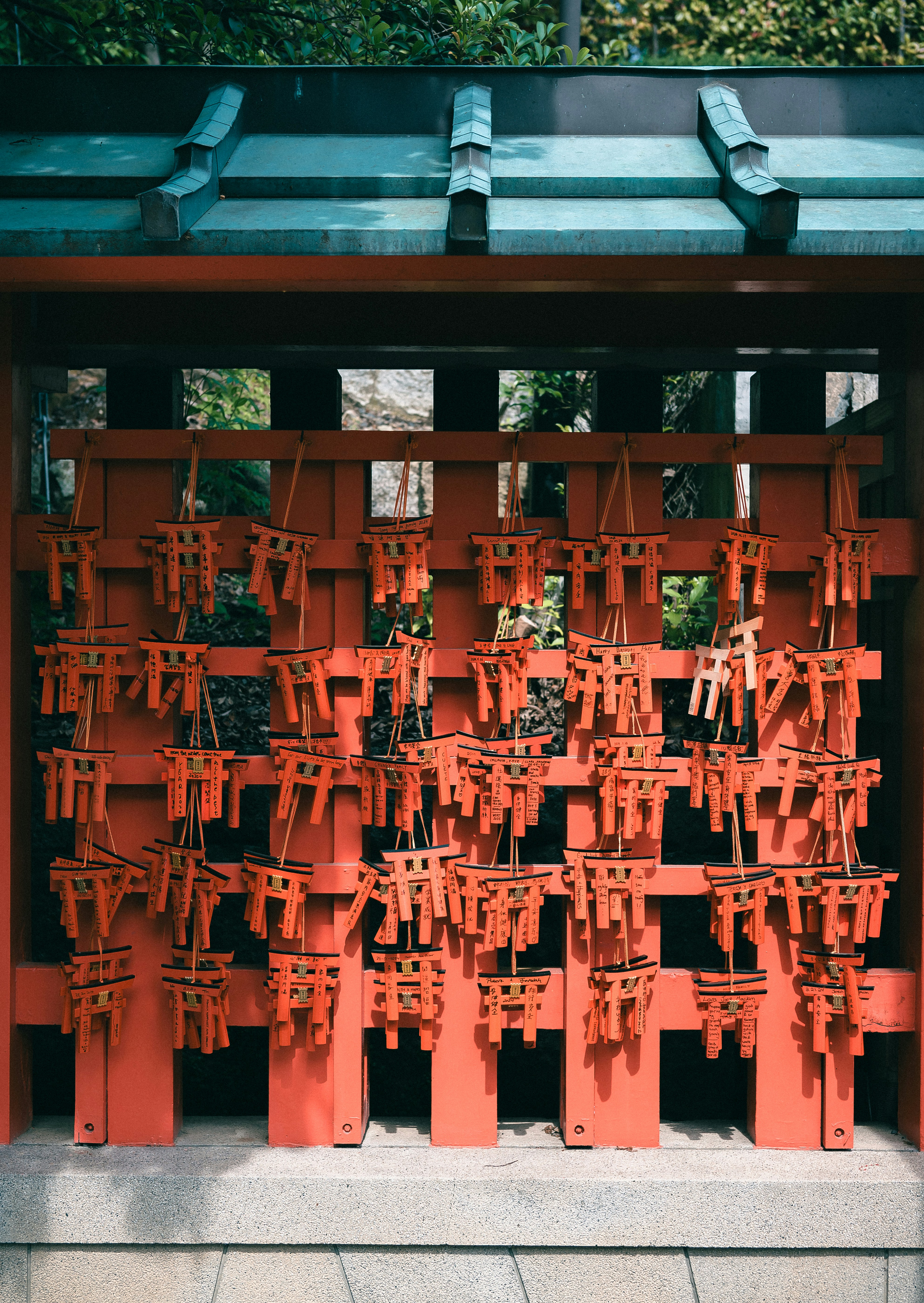 Dense grid of red ema plaques arranged in rows in front of a teal shrine roof, with a stone base at the bottom. The repetitive geometry and bold color contrast highlight traditional shrine elements.