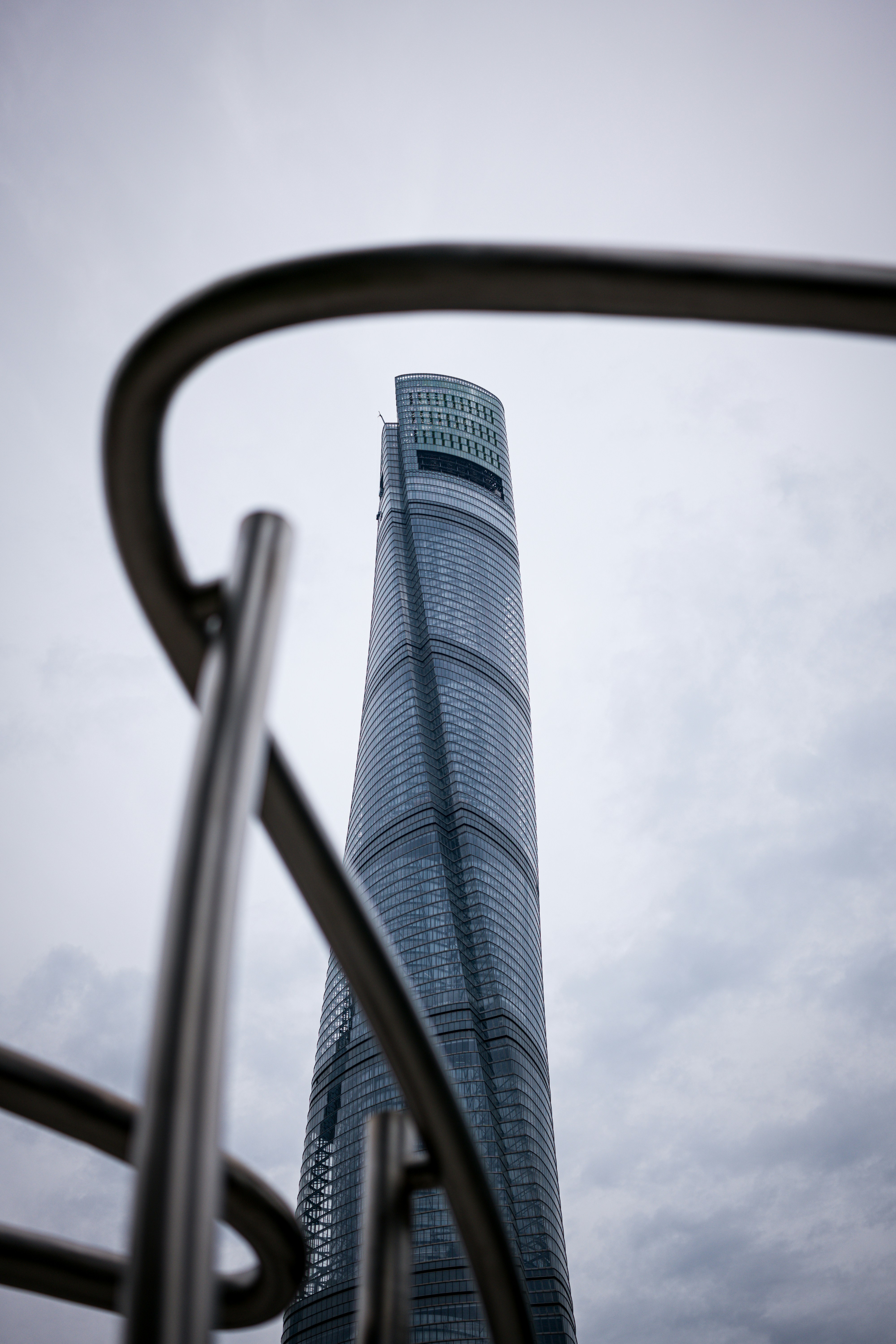 Tall glass skyscraper rises through curved metal rails in the foreground, captured from a low, upward angle. The composition emphasizes architectural scale against a cloudy sky.