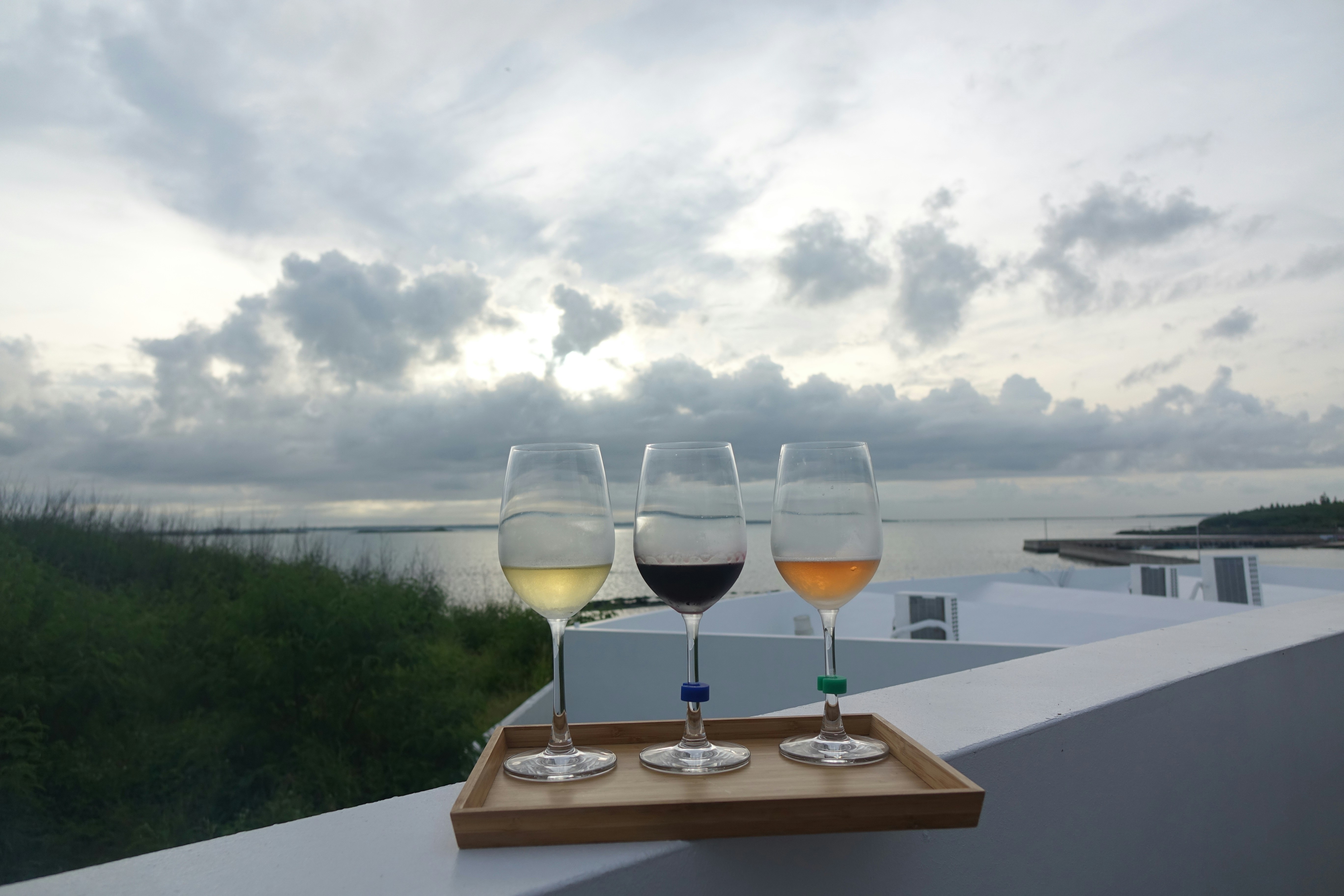 Three wine glasses on a wooden tray sit on a balcony railing, overlooking the sea as dramatic clouds roll in at sunset.