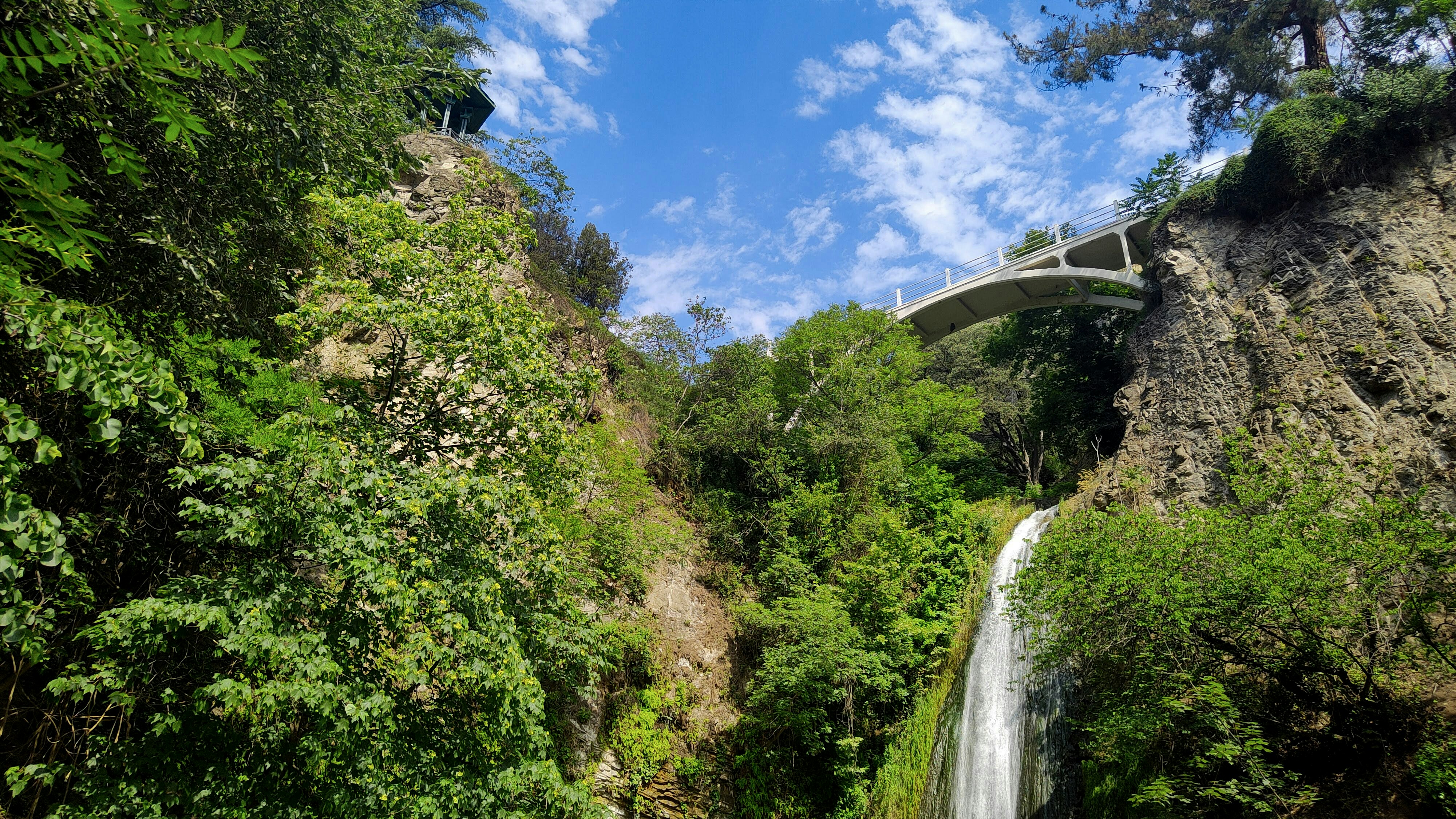 Waterfall cascades down a rocky canyon framed by lush trees, with a white bridge arching overhead under a bright blue sky.
