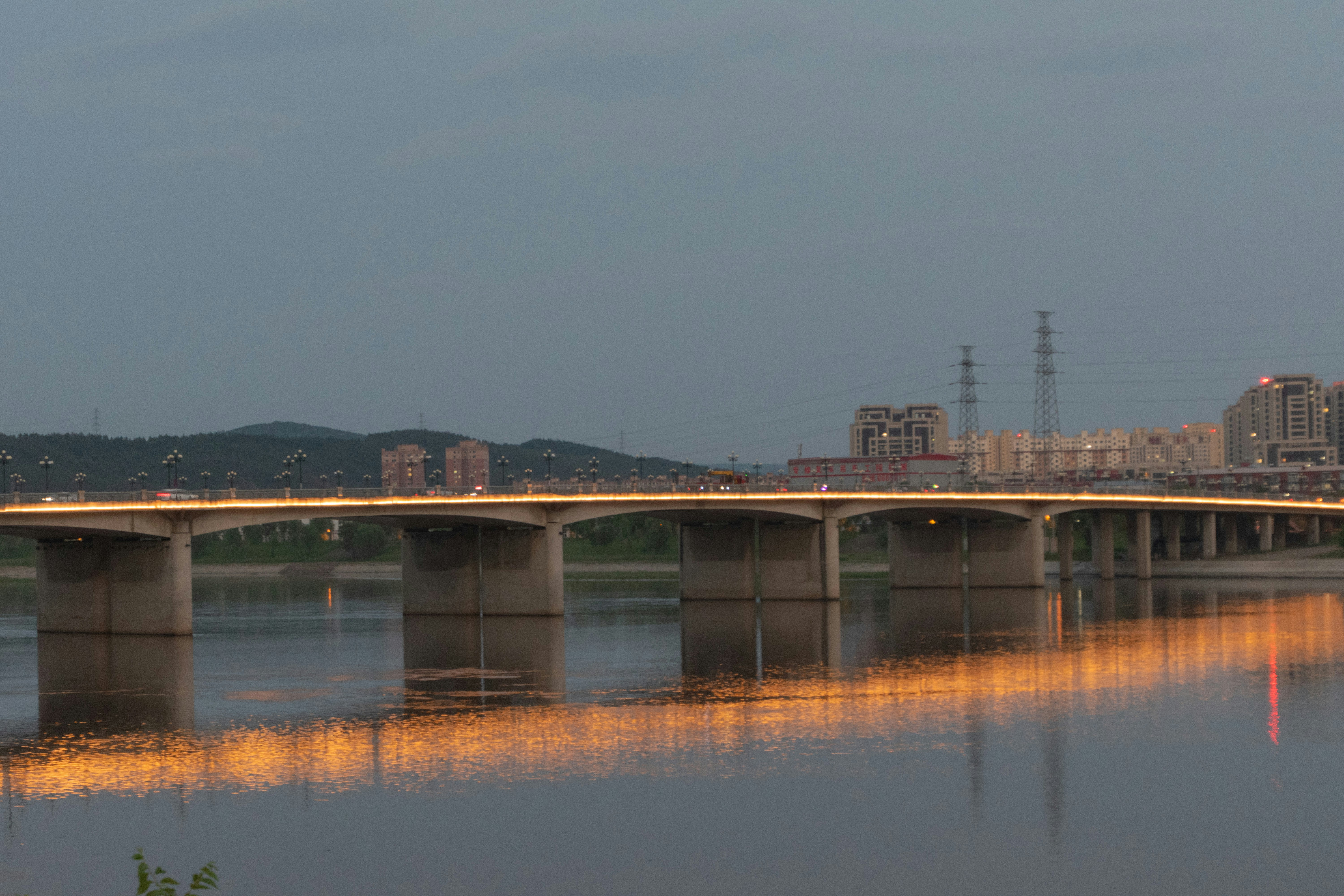 A bridge gracefully spans a river, illuminated by soft lights that reflect on the water's surface, with a backdrop of urban architecture and rolling hills.