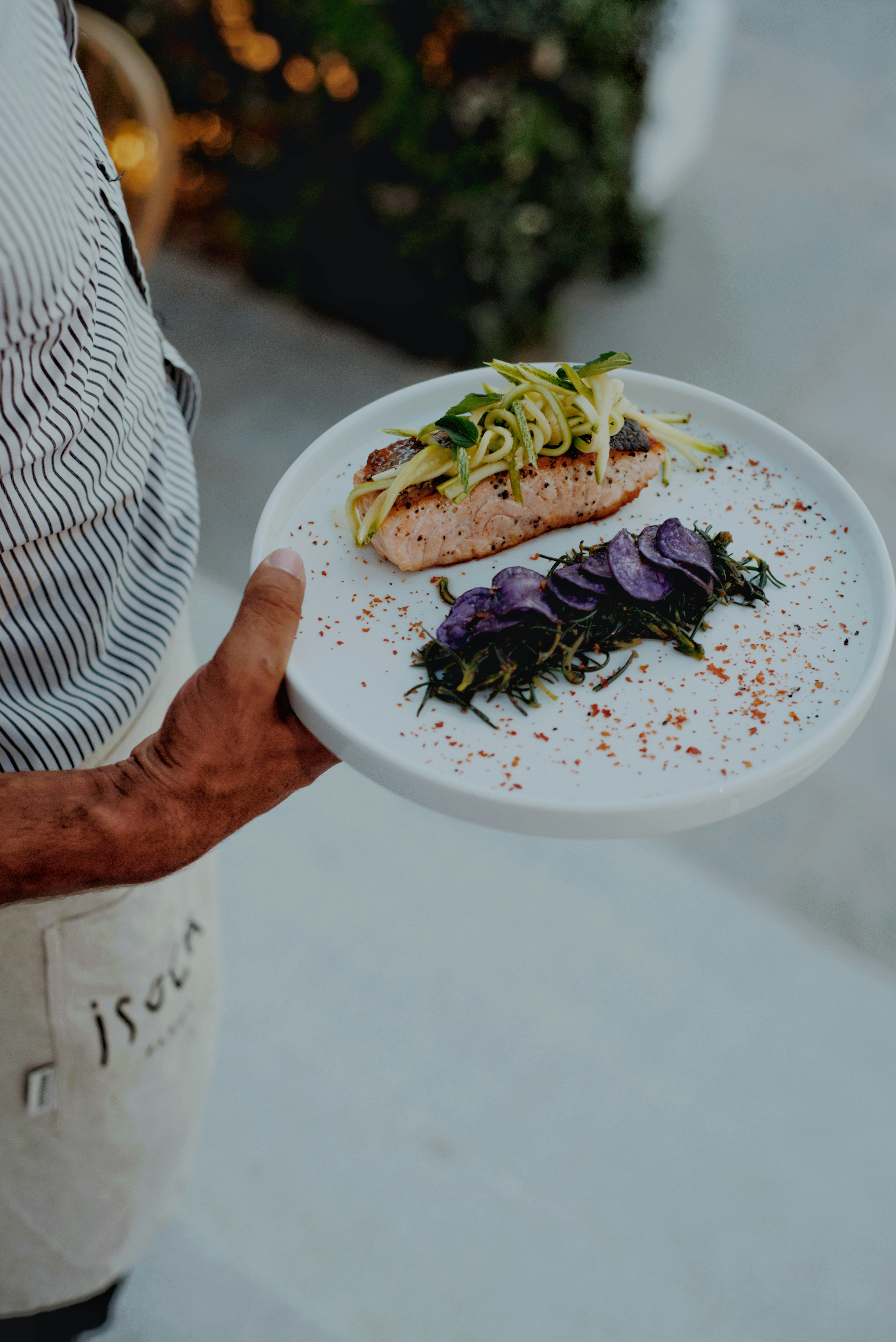 Photograph of a salmon fillet topped with fennel and herbs on a white round plate. A bed of greens and purple potato crisps sits beside the fish, held by a person wearing a striped shirt.