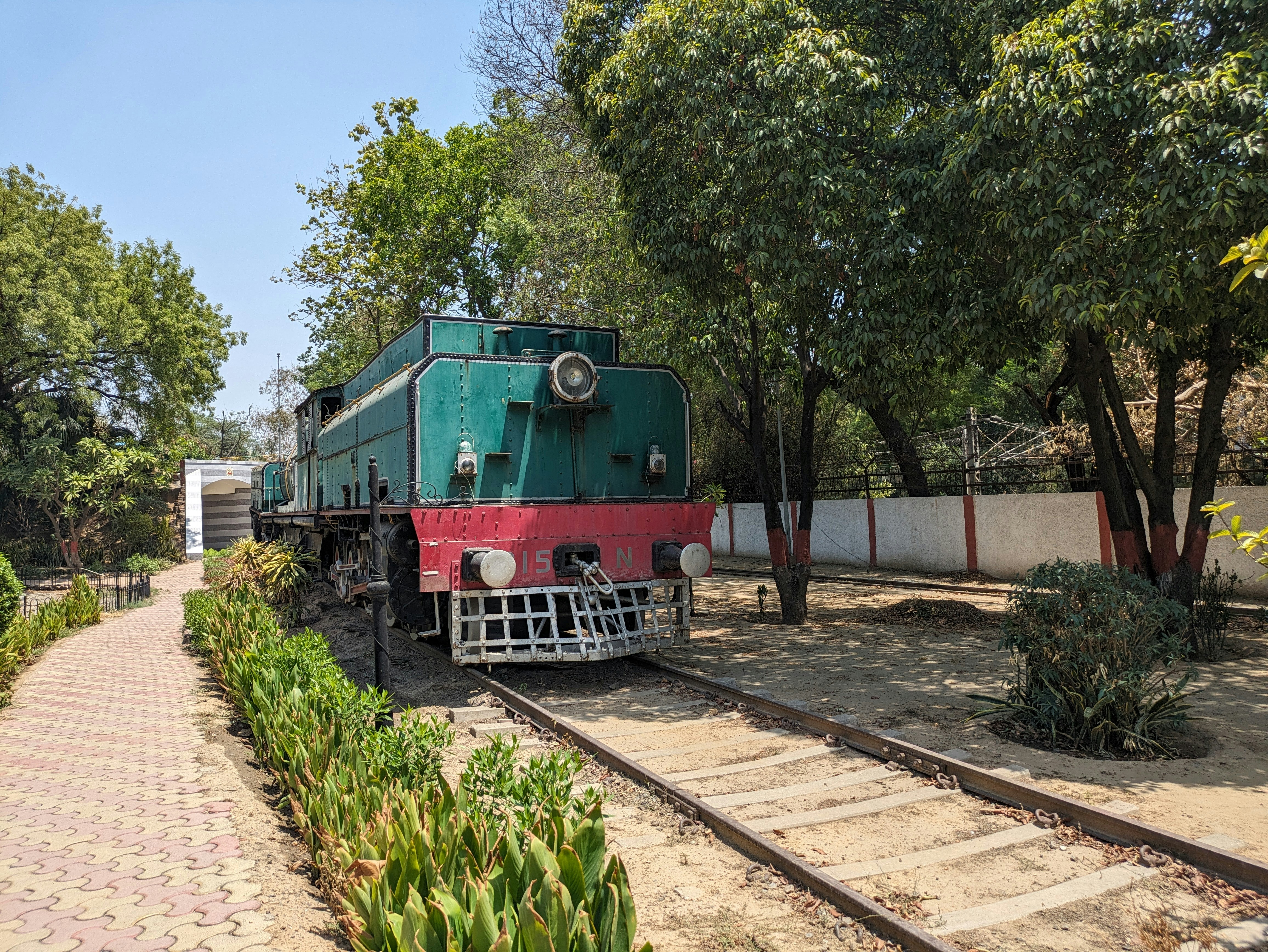 A vintage green locomotive rests on rusted tracks beside a tree-lined park, with a brick-paved path running along the left. This photograph highlights a heritage rail exhibit in a tranquil outdoor setting.