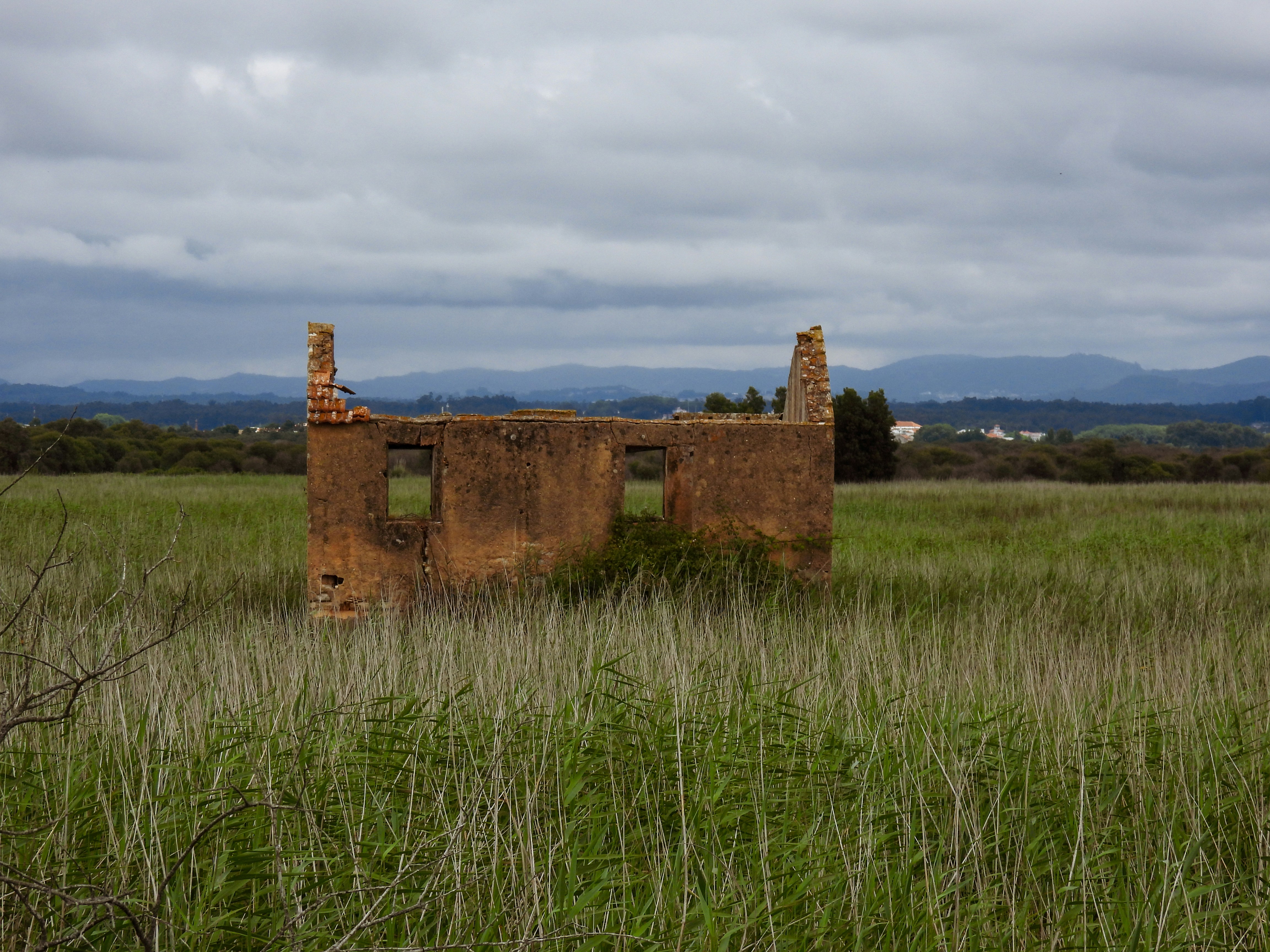 Photograph of a small brick ruin in a grassy field with distant mountains and a cloudy sky.