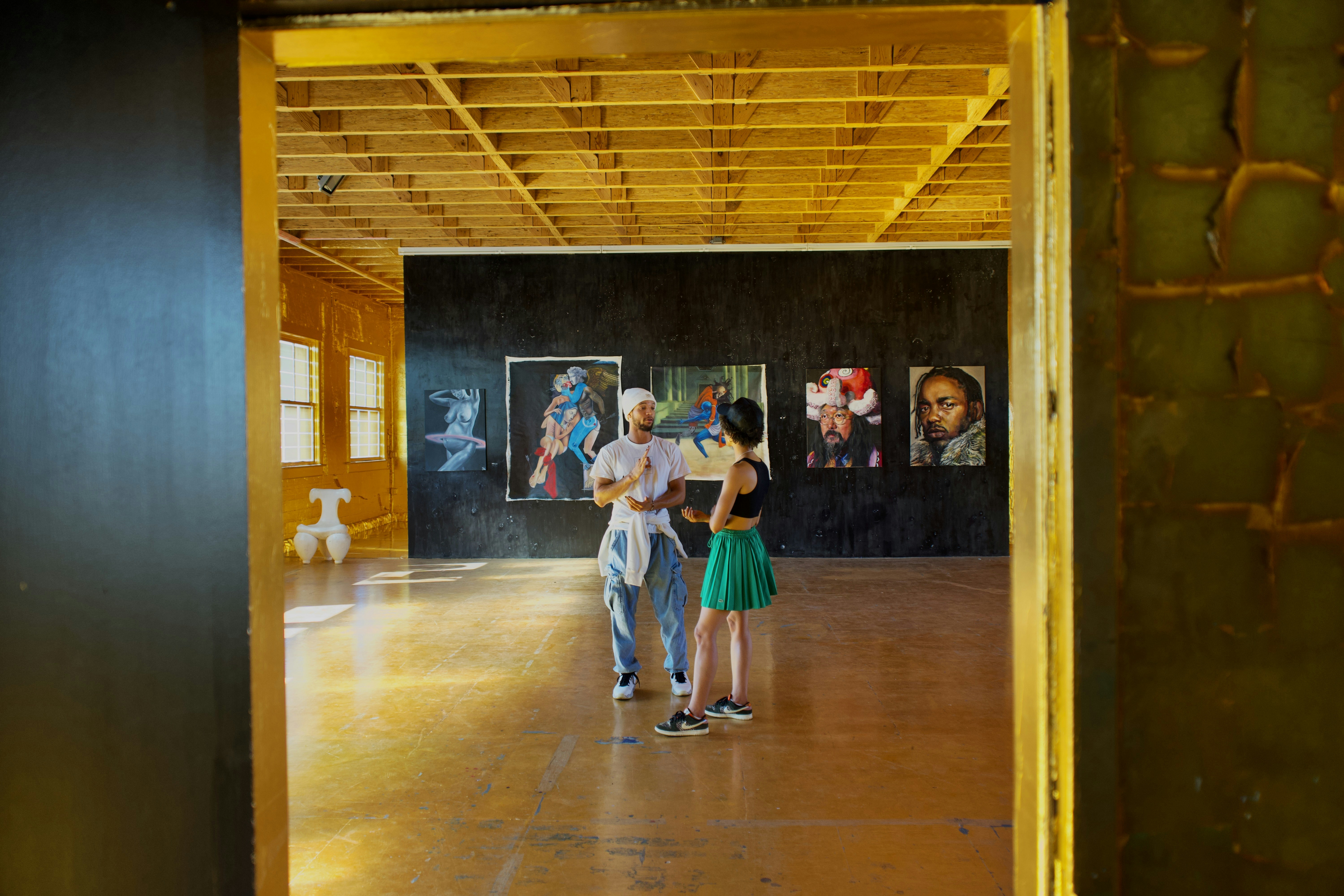 Two people stand engaged in conversation in a warmly lit gallery, surrounded by portraits on dark walls.