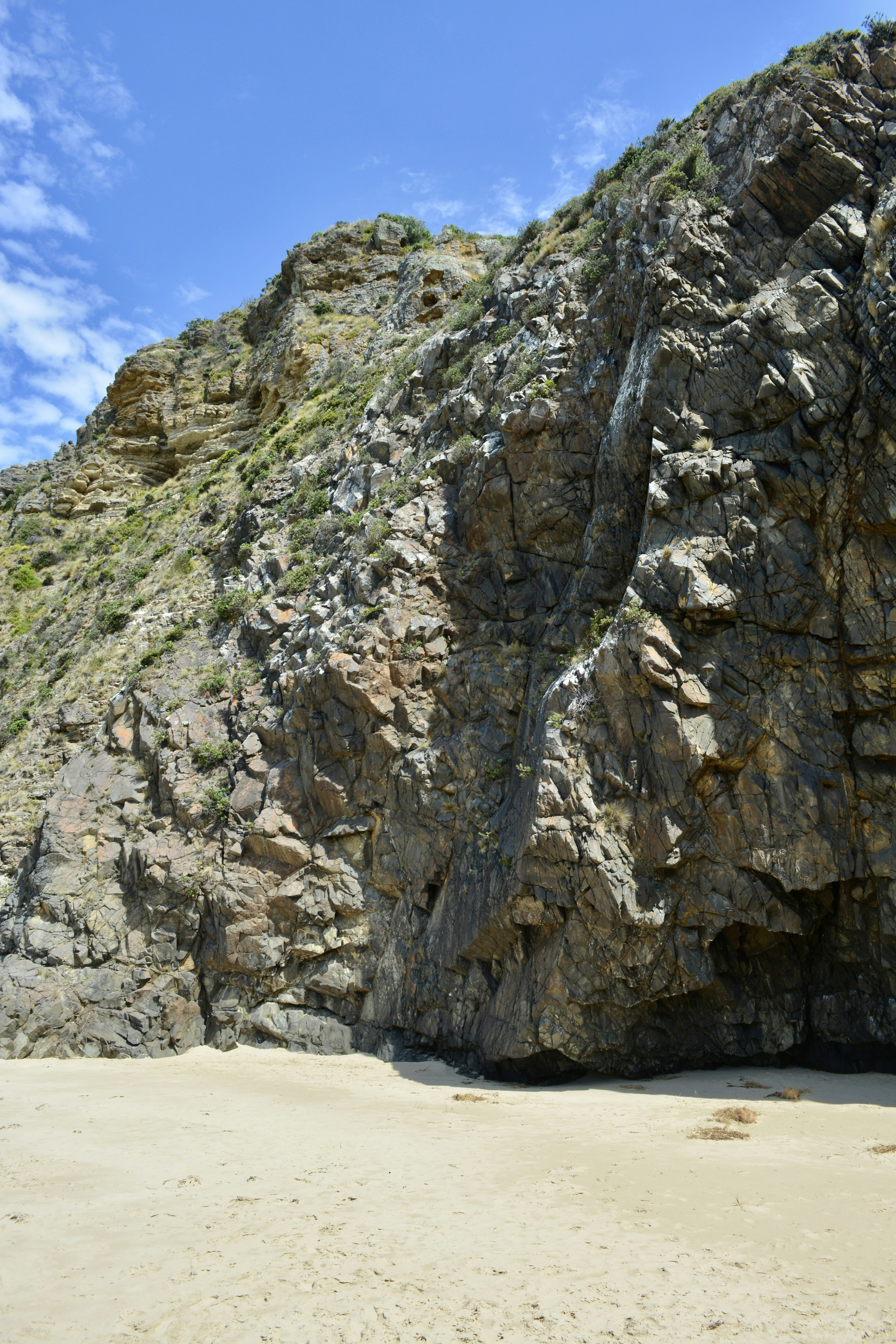 Jagged cliffs rise dramatically against a clear blue sky, showcasing layers of rock and vegetation along a sandy beach.
