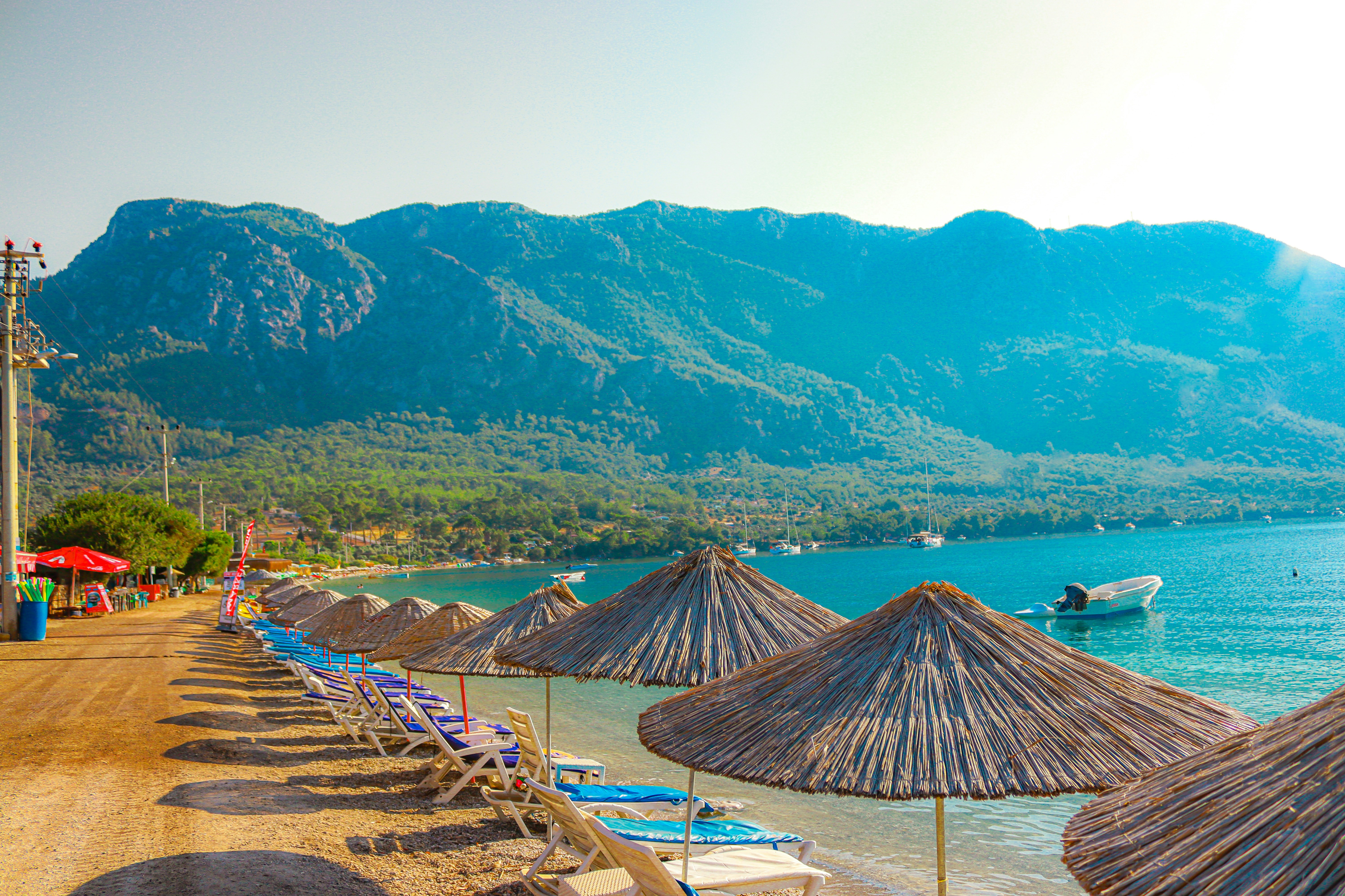 Lounge chairs lined along a sandy beach with straw umbrellas, framed by lush mountains and a clear blue sea. Perfect for a tranquil getaway.