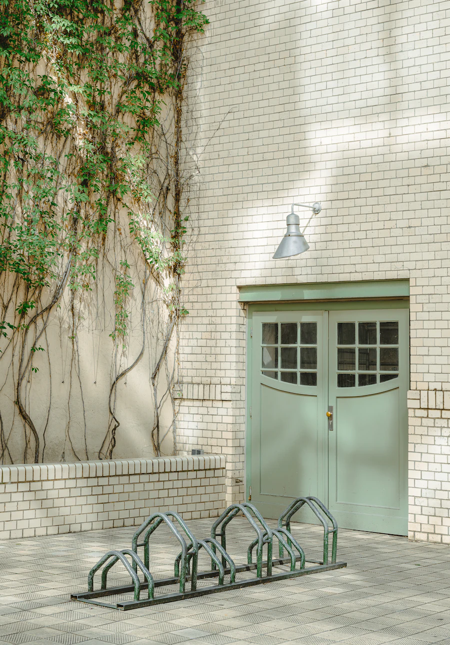 Mint green double doors set in a pale brick courtyard wall, with climbing ivy on the left and a metal bike rack in the foreground. Soft daylight creates gentle shadows across the facade.