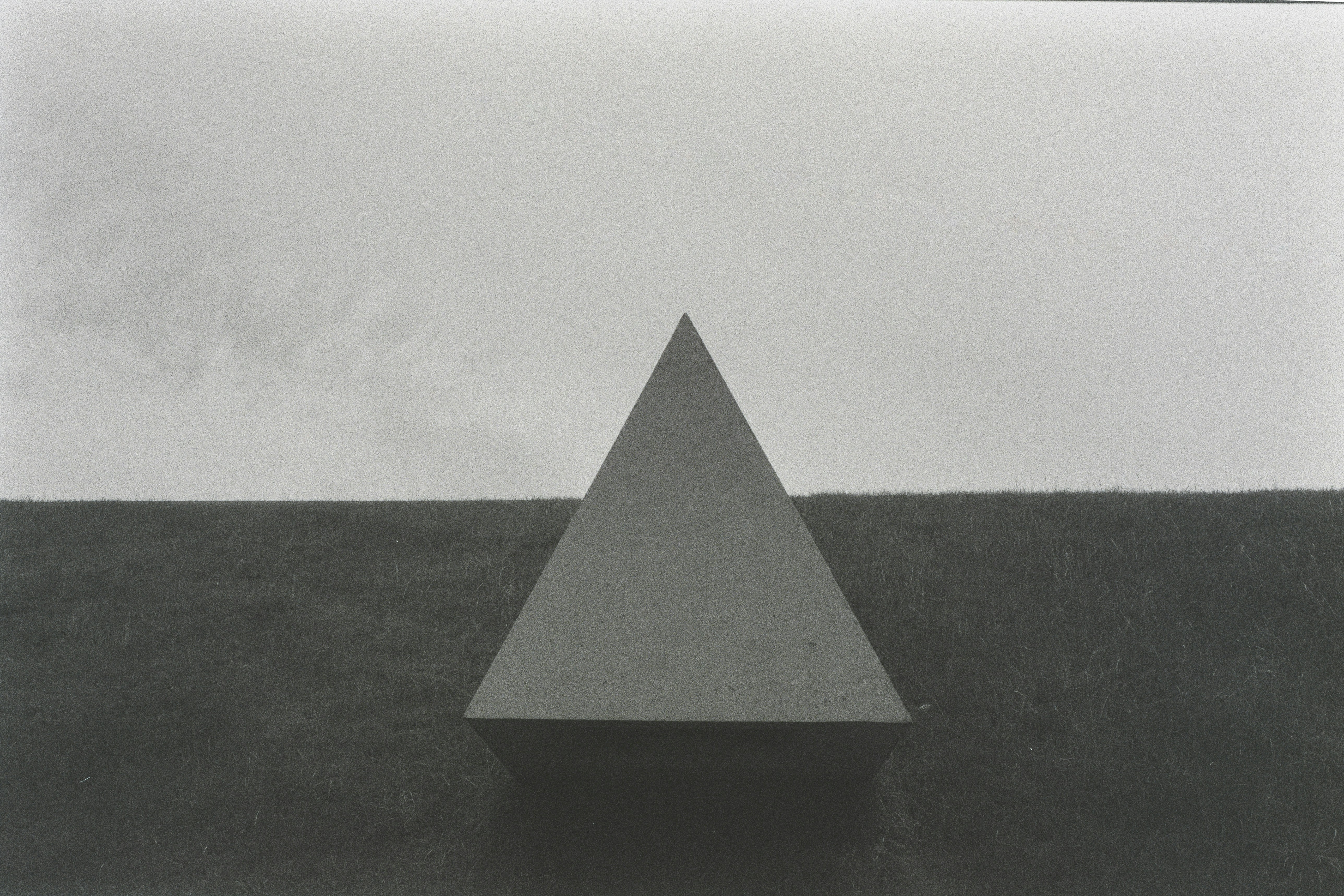 Grayscale photograph of a solitary white triangular prism on a dark surface with a cloudy sky in the background.