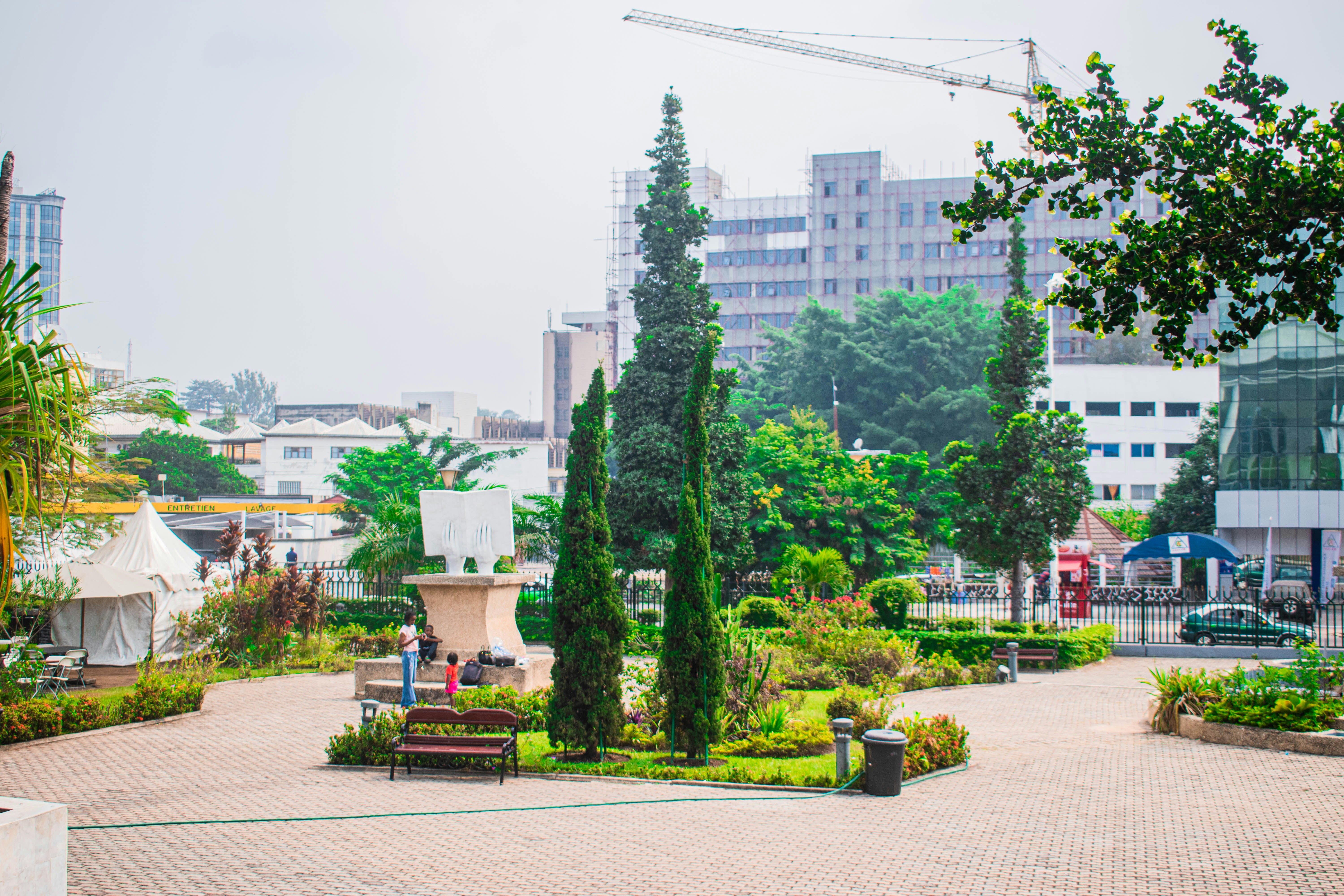 Lush greenery and neatly arranged gardens create a serene space in the heart of a bustling city, with tall trees and a decorative fountain at the center.
