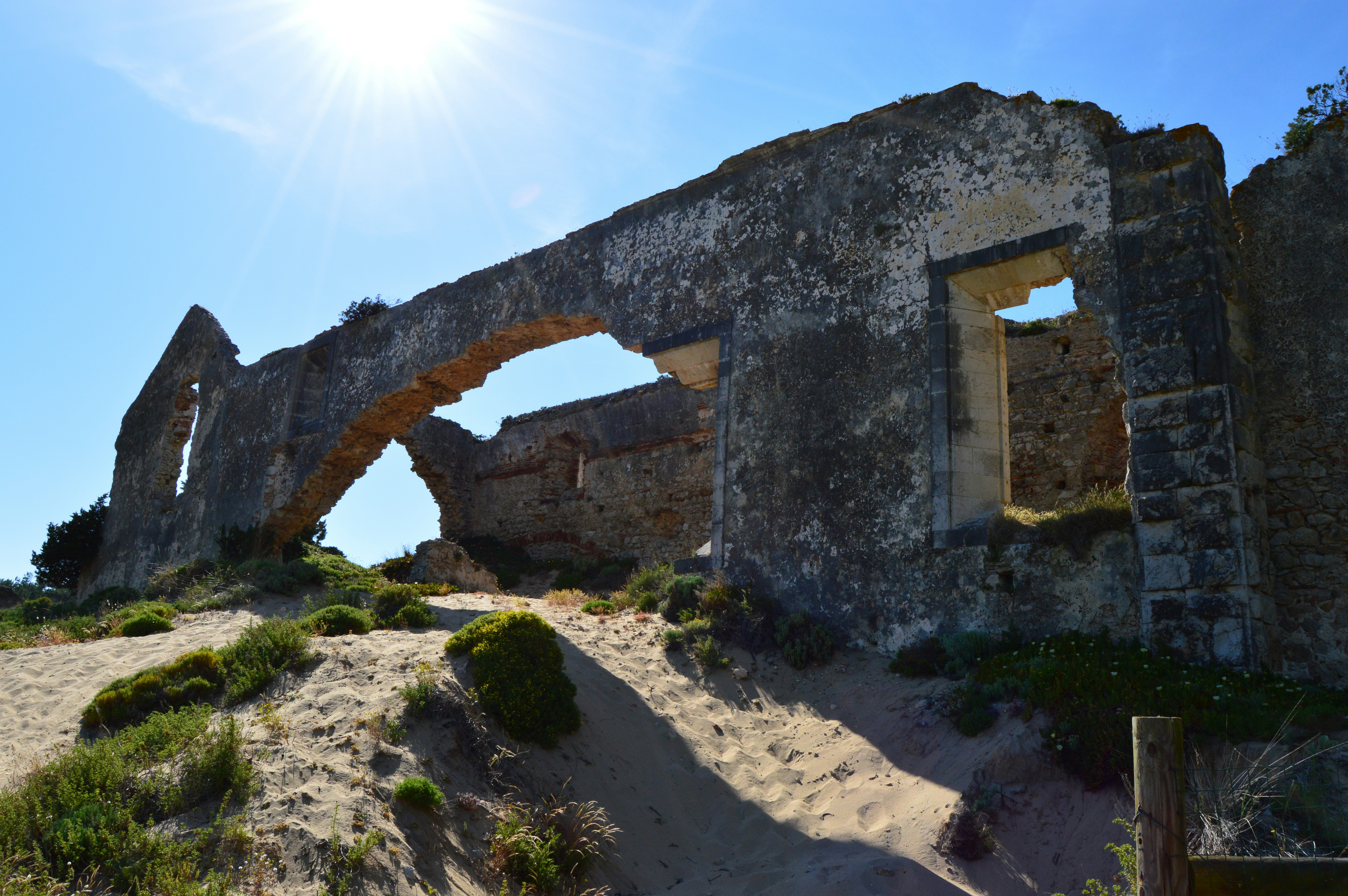 Ancient stone ruins with arches and windows under a bright sun, casting shadows on sandy ground.