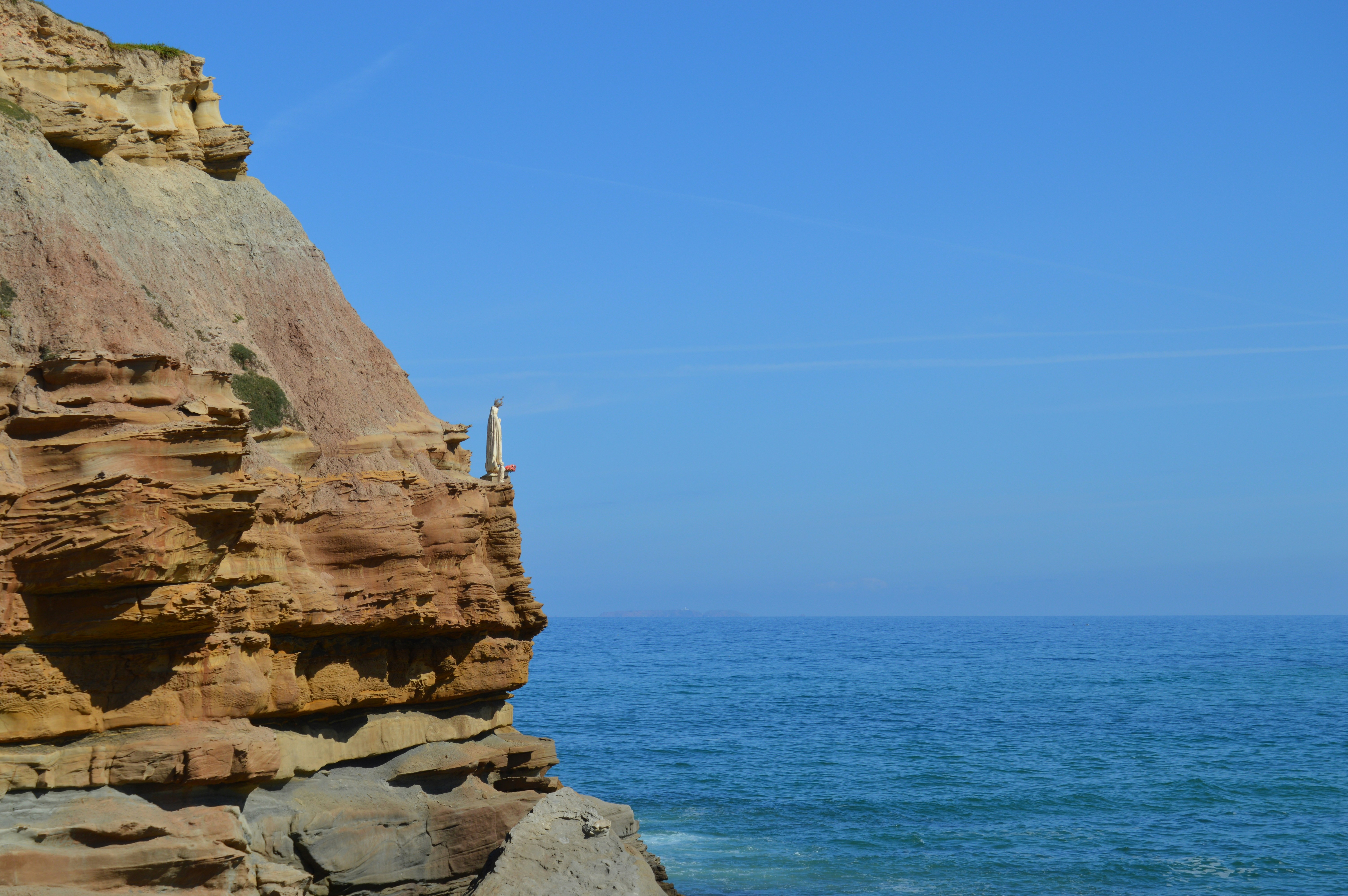 Person standing atop a rugged cliff overlooking a vast blue ocean under a clear sky.