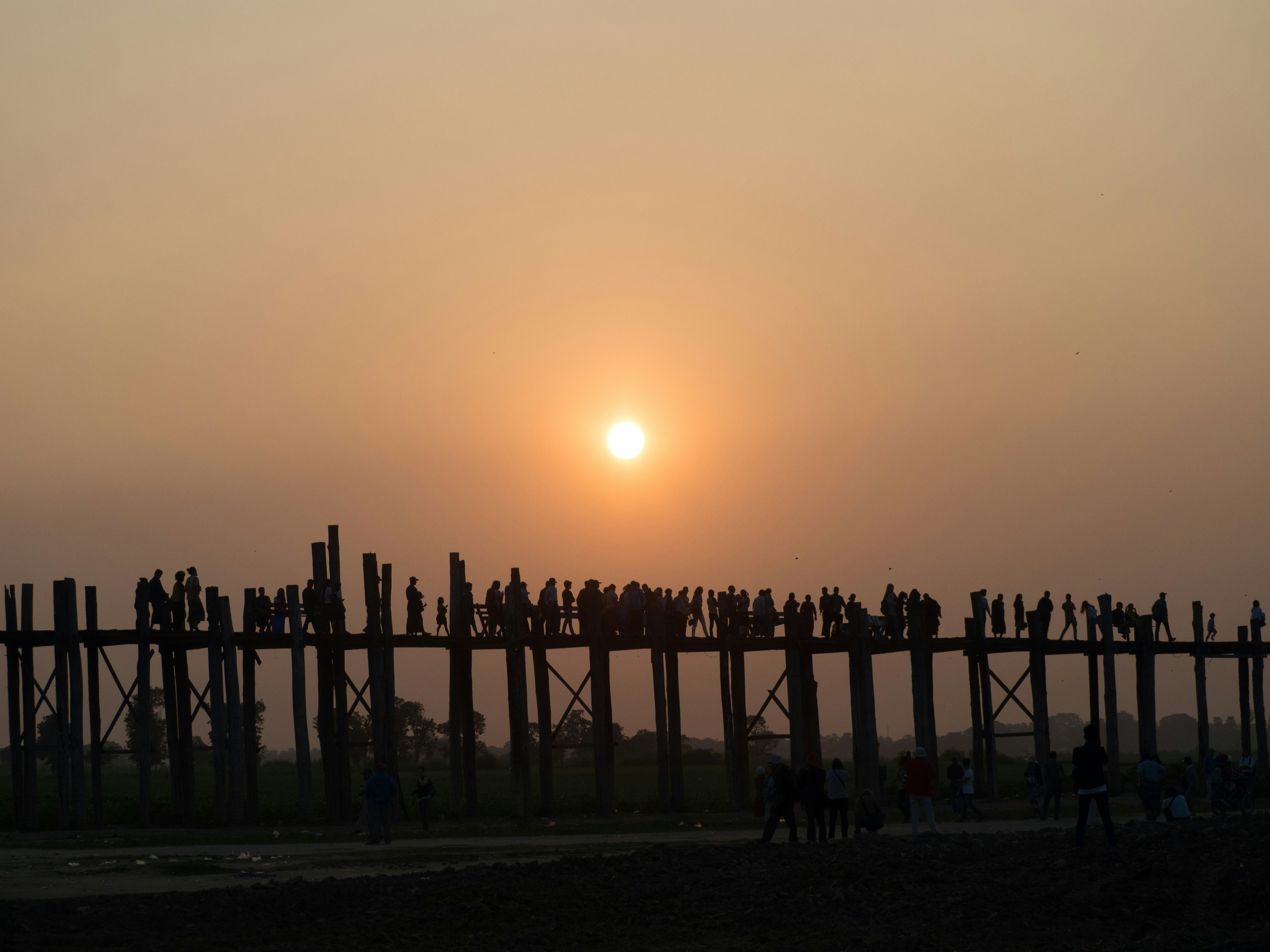 Silhouetted figures stand atop a long wooden bridge against a hazy sunset sky.