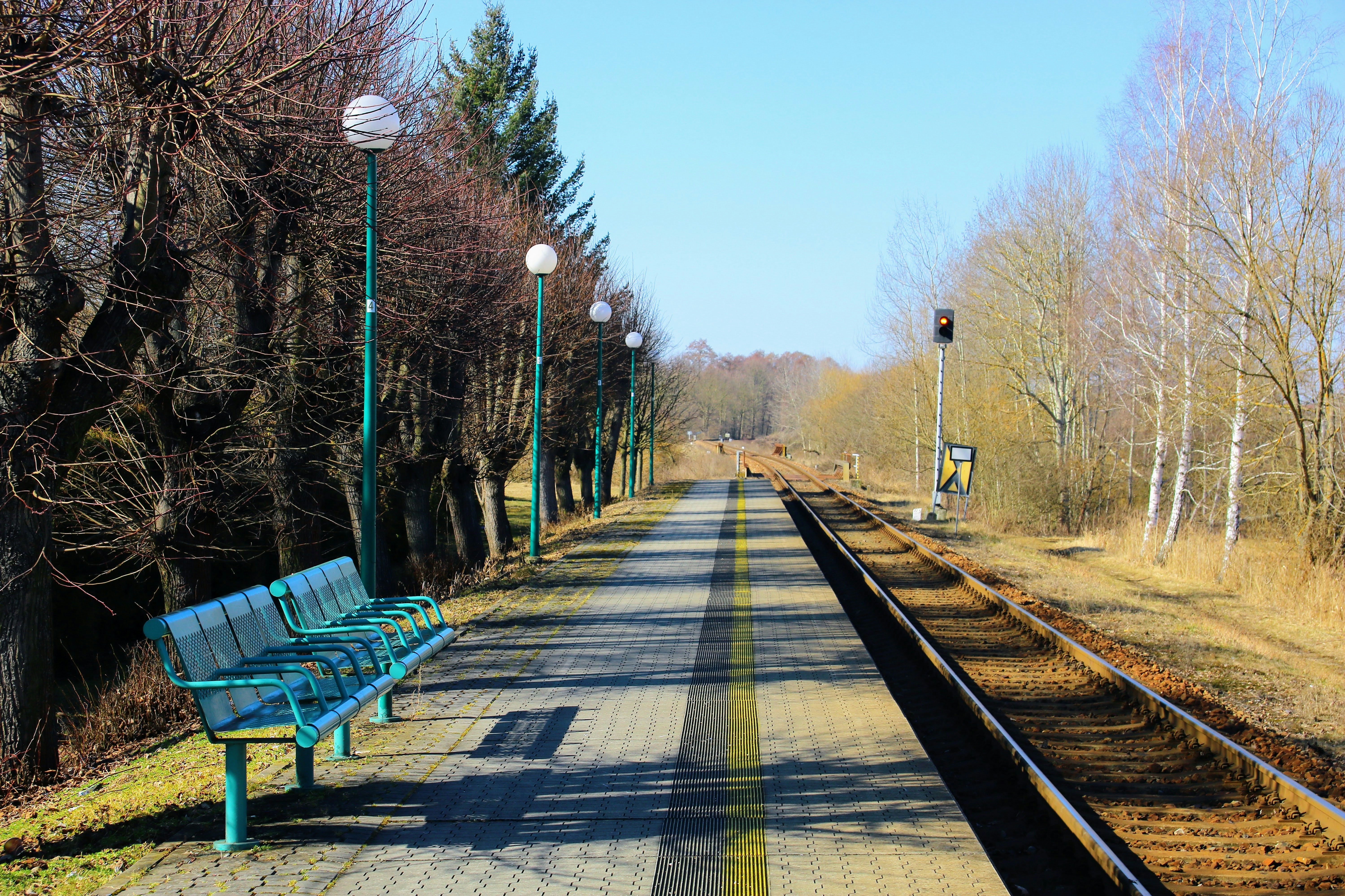 Sunlit train station platform with benches, lamp posts, and bare trees lining the tracks.