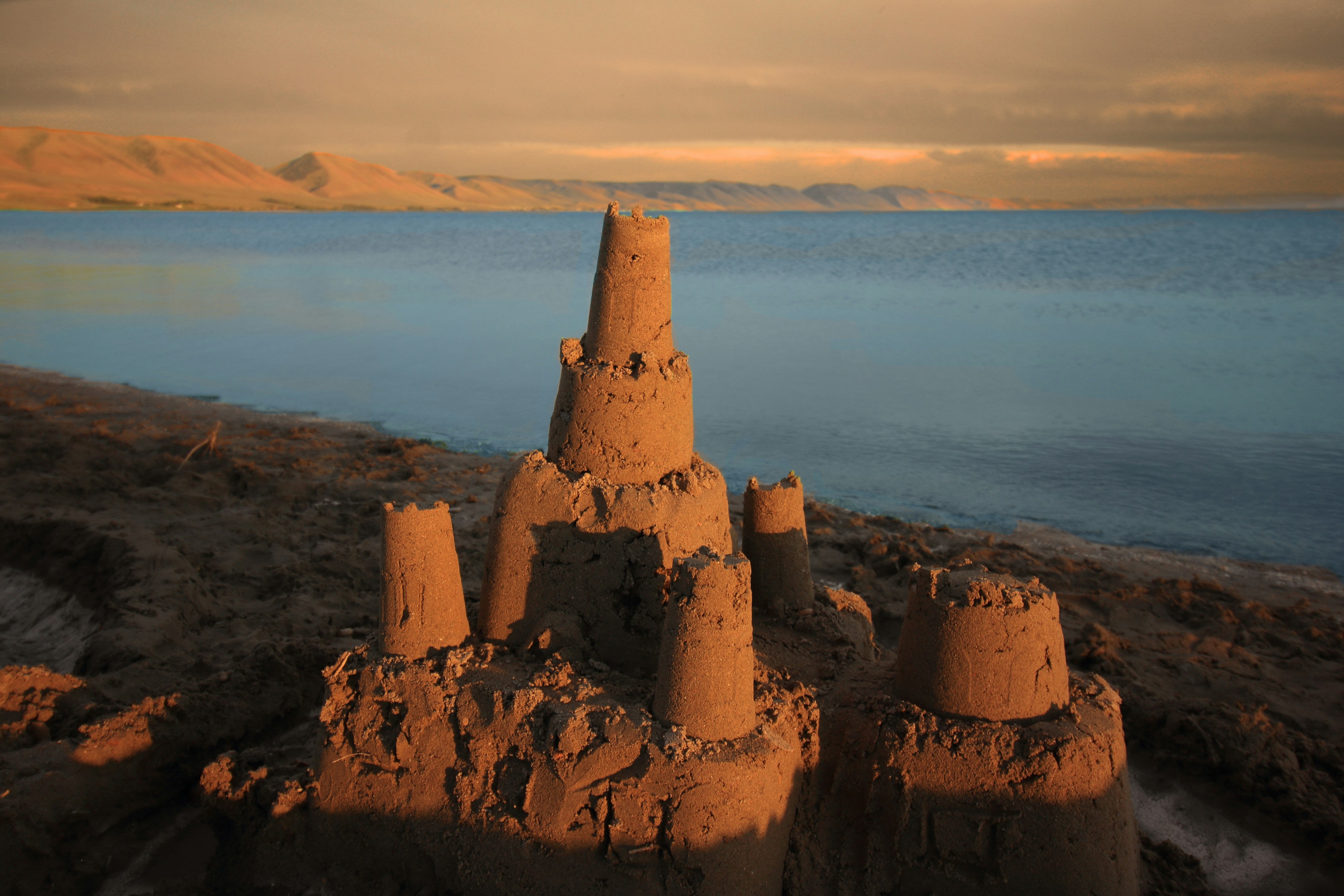 Sandcastle on a beach with a serene lake and sunset in the background.