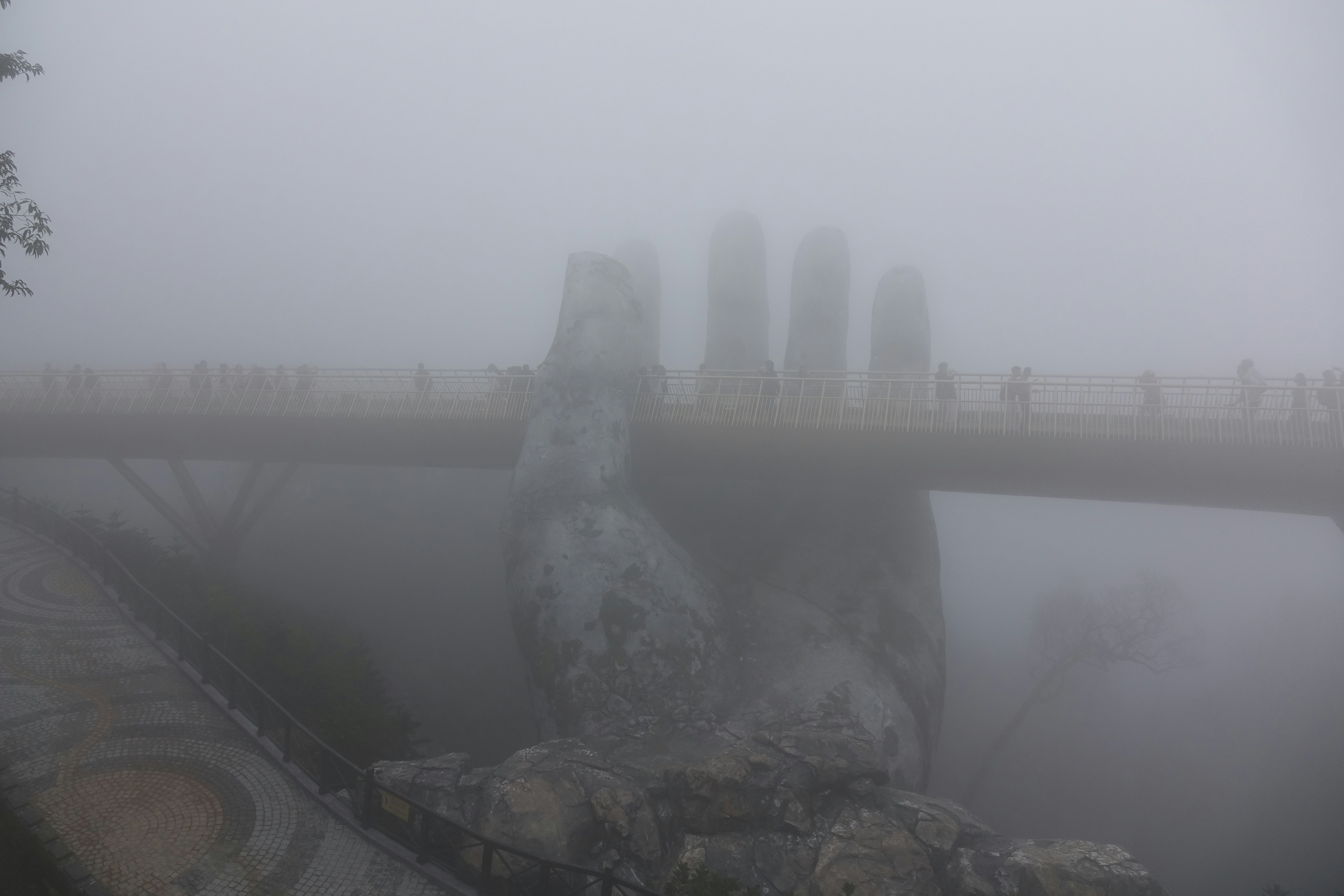 A colossal stone hand rises from the rocky base beside a pedestrian bridge shrouded in dense fog.