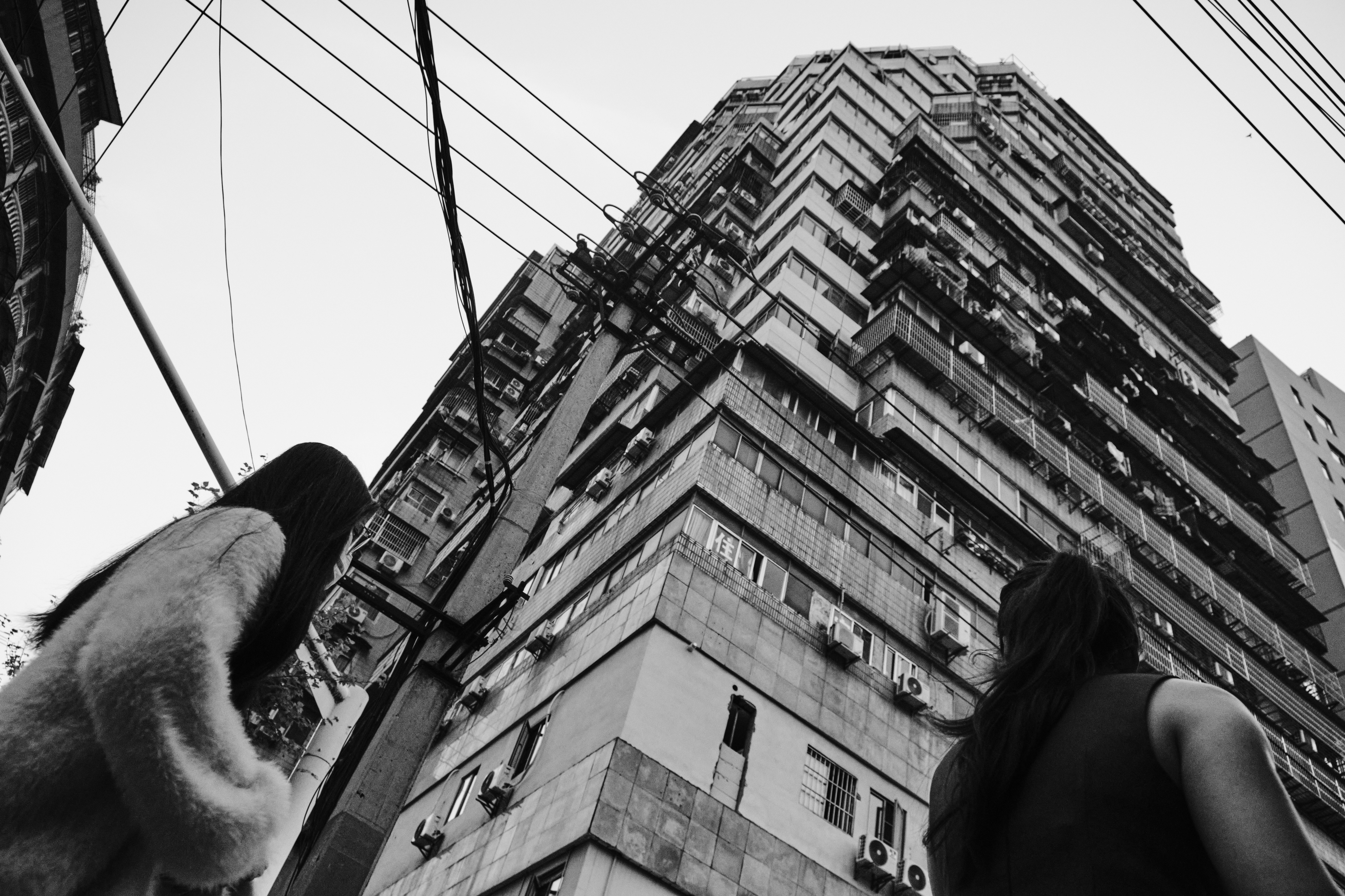 Two people gaze up at a towering high-rise building under a network of overhead cables.