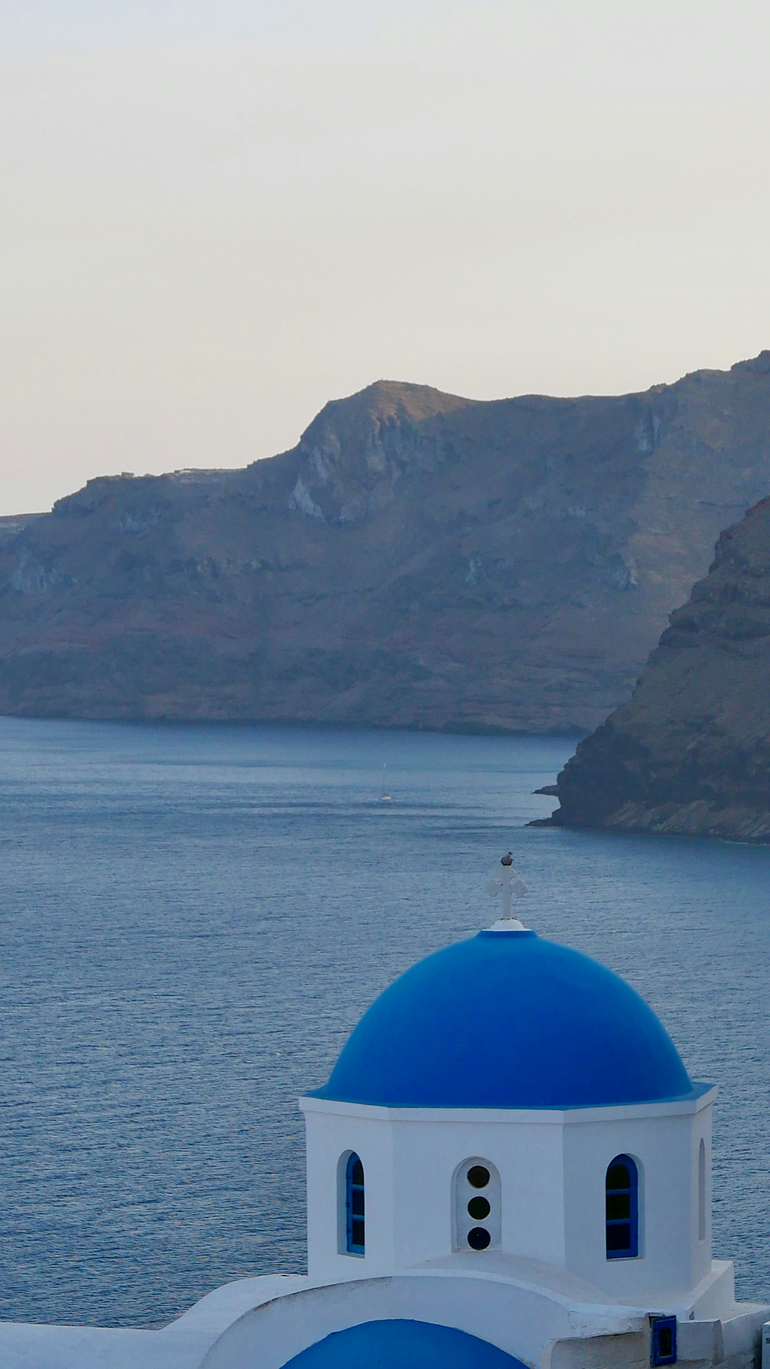 Blue-domed chapel sits at the cliff foreground with calm sea stretching toward rugged coast. The scene emphasizes the iconic architecture against the rocky backdrop.