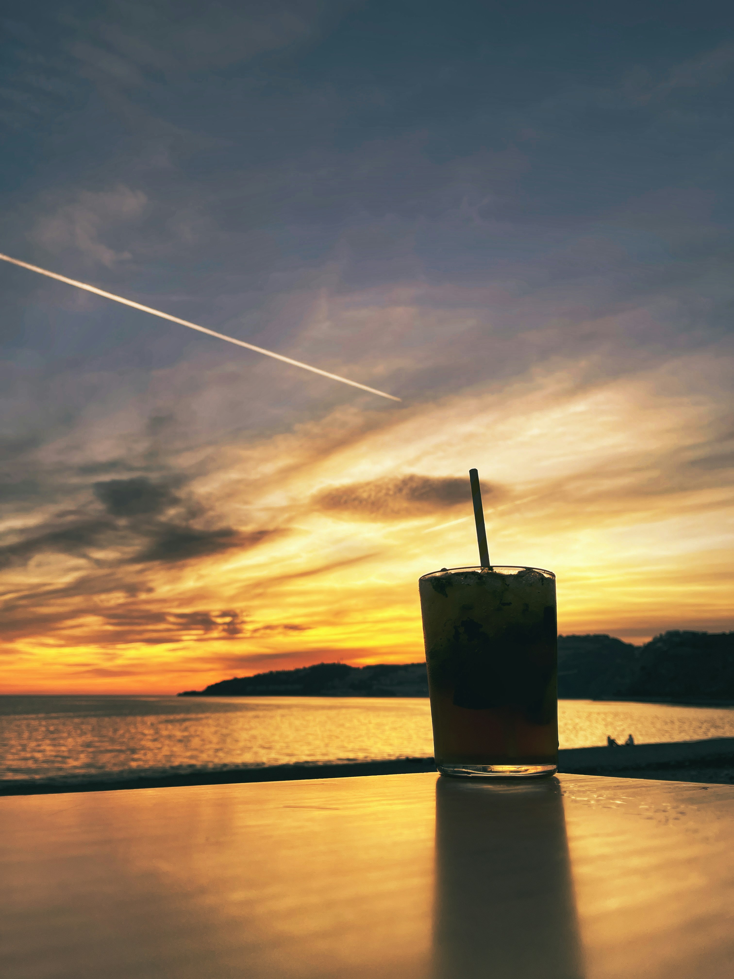 A refreshing cocktail rests on a table as the sun sets over the horizon, painting the sky with vibrant hues. The scene captures the essence of relaxation and evening leisure.