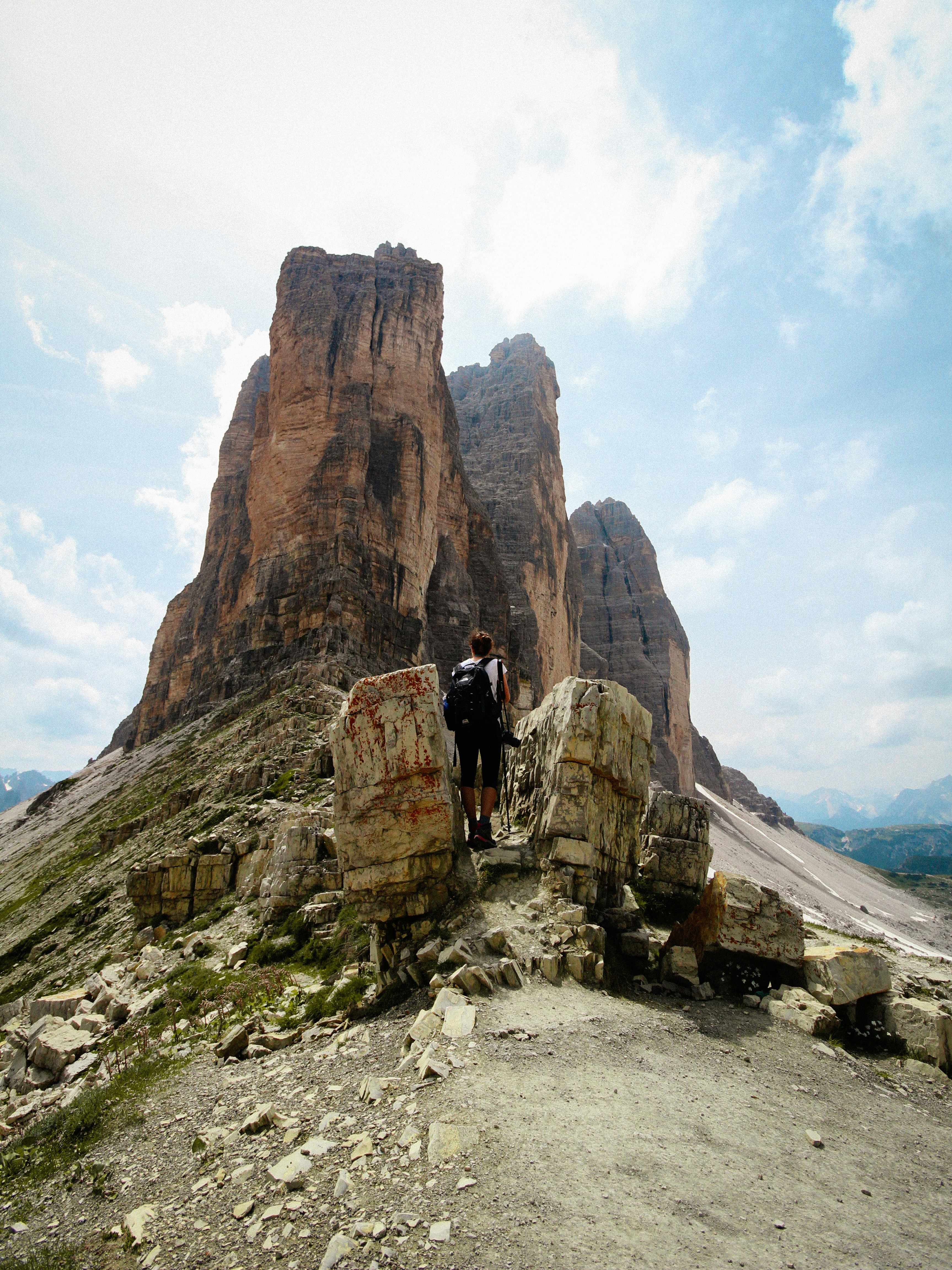 A hiker stands atop a rocky outcrop, gazing at towering cliffs under a partly cloudy sky. The rugged landscape showcases the majesty of nature's formations.