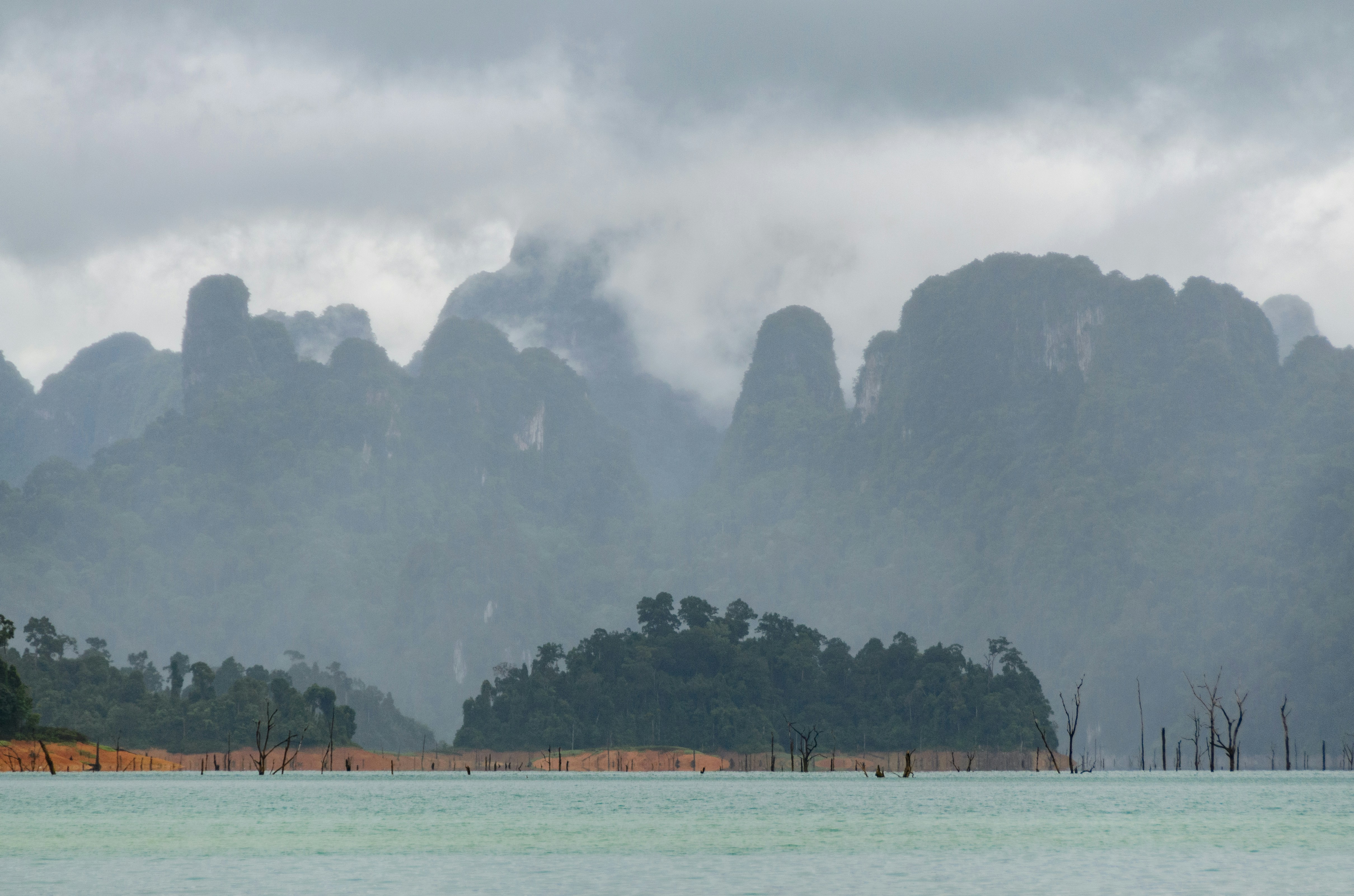 Tranquil lake with mist-shrouded mountains in the background under a cloudy sky.