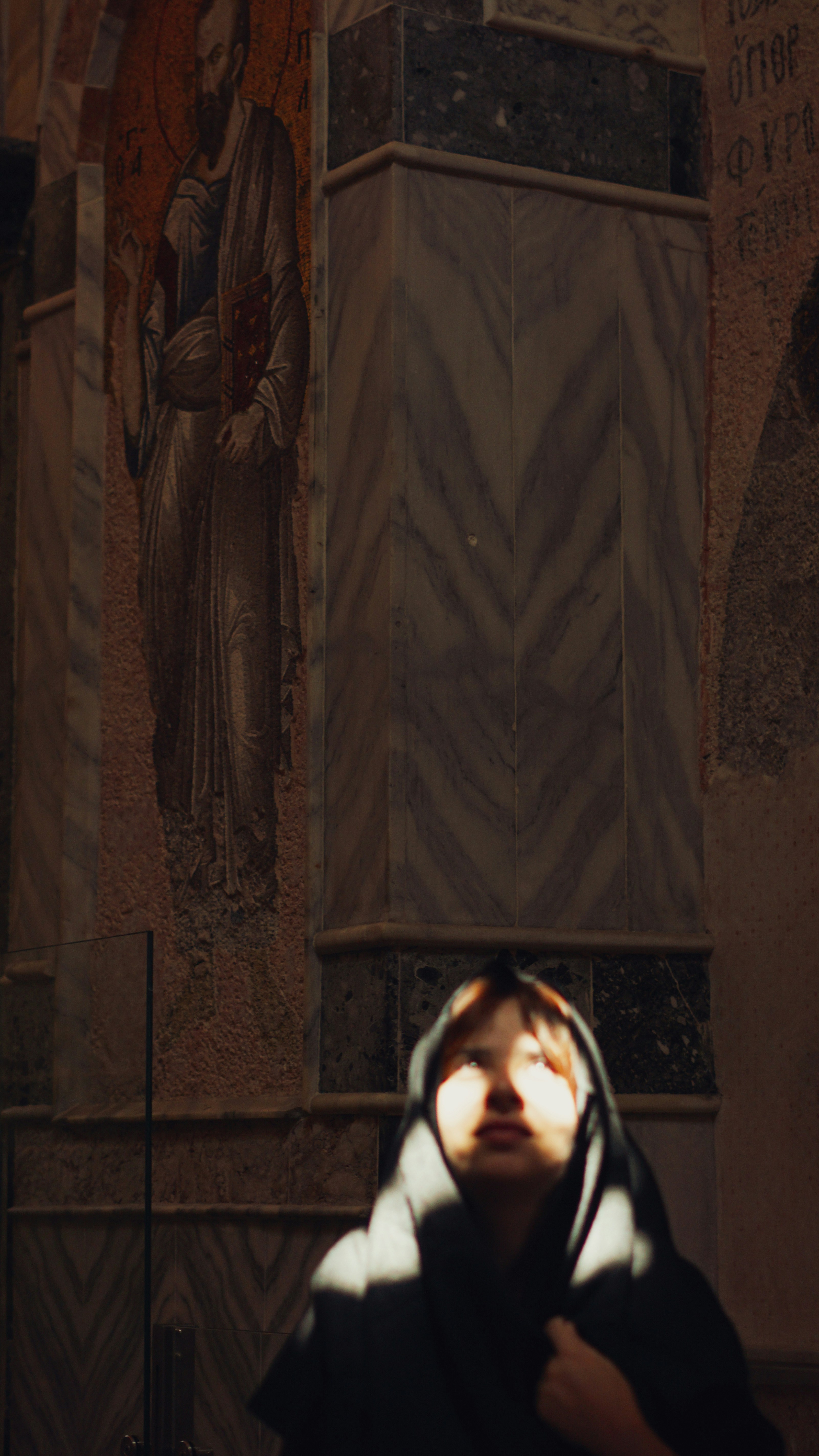 Low-Light Reverie: Hooded Figure Before a Saintly Mural