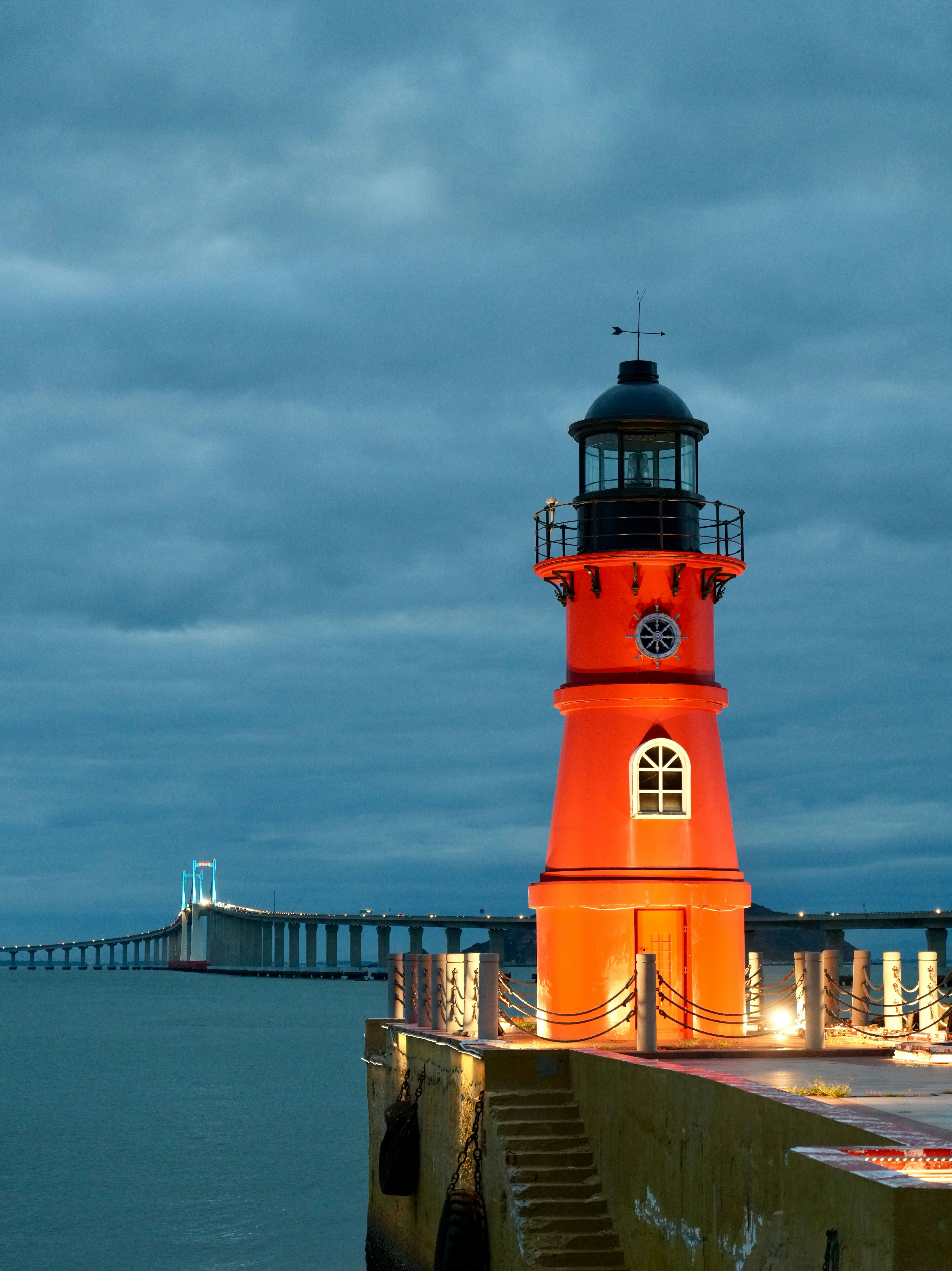 Vibrant orange lighthouse stands tall against a moody sky, guiding vessels through twilight. A distant bridge looms in the background.