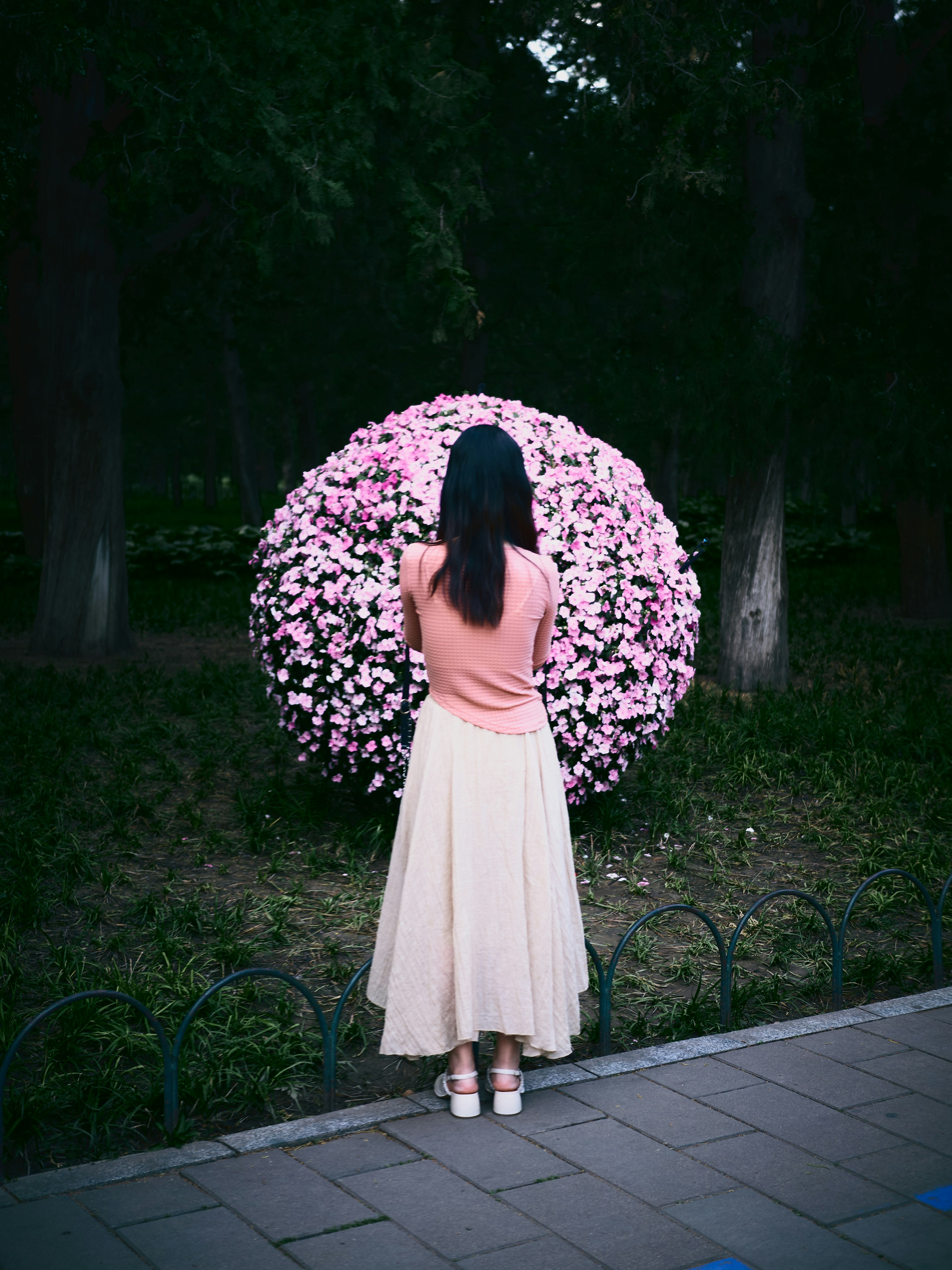 A photograph of a woman with dark hair standing with her back to the camera, facing a large pink flower sphere in a park. The scene emphasizes pastel color and a serene, contemplative mood.
