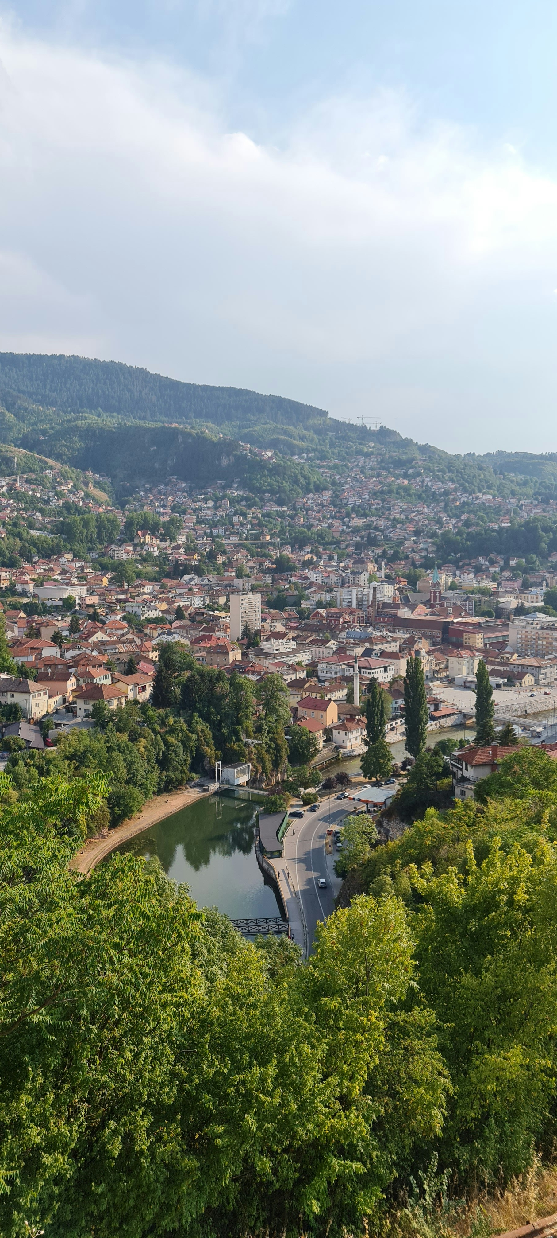 Panoramic cityscape with red-tiled rooftops and a winding river bordered by trees, set against green hills under a blue sky.