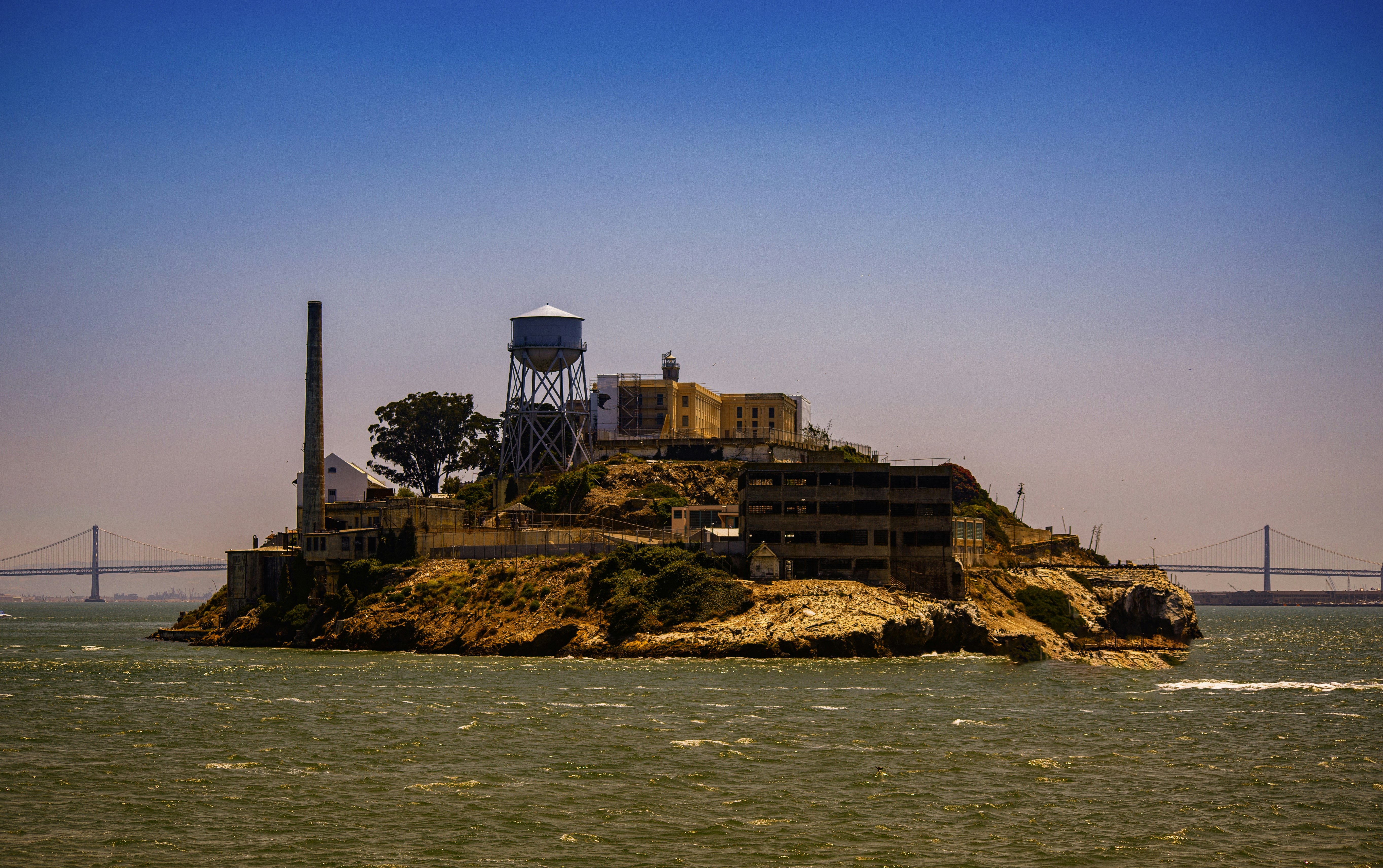 Alcatraz Island with its historic buildings and water tower, surrounded by choppy waters and distant bridges.