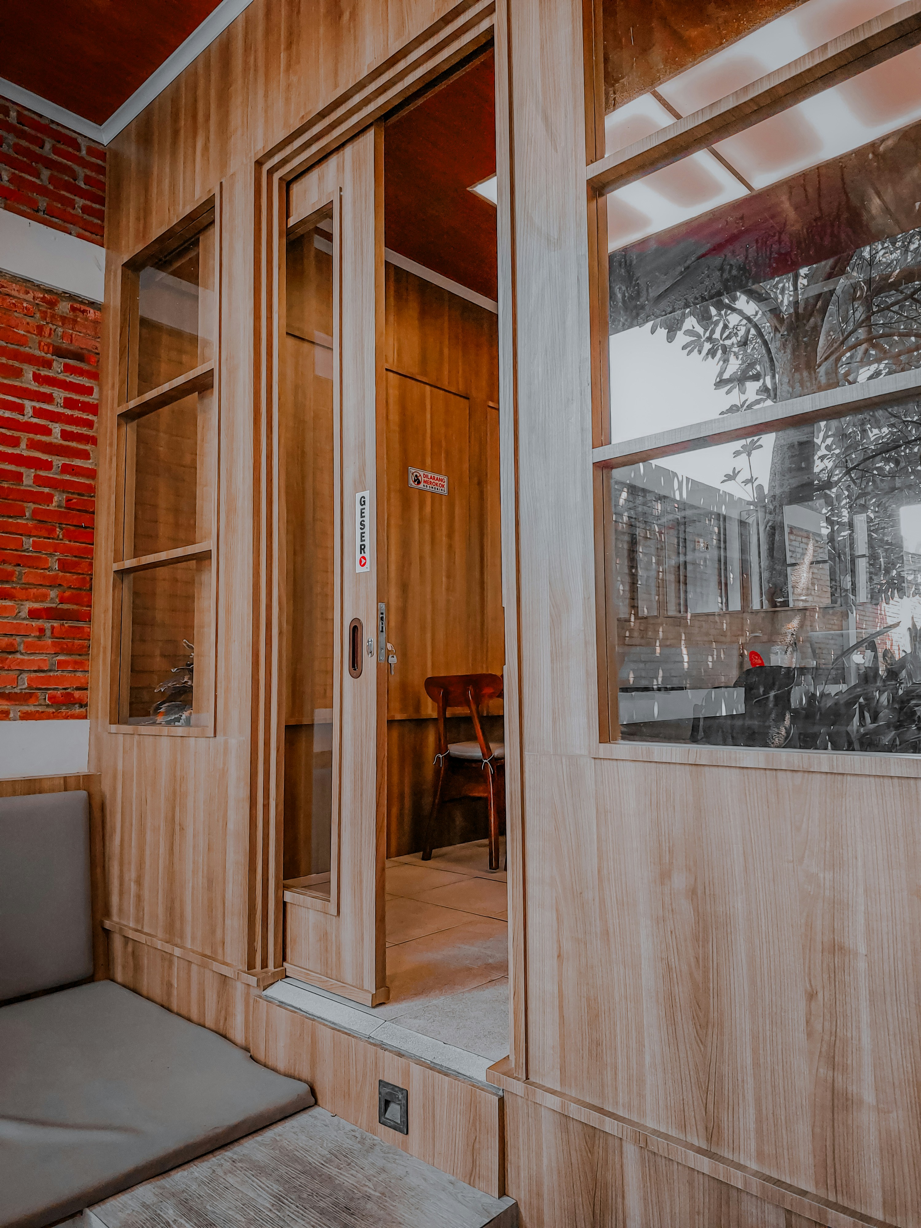 Wooden doorway leading into a serene space with a chair visible inside, framed by warm tones and rustic brick. A hint of greenery is seen through the glass.