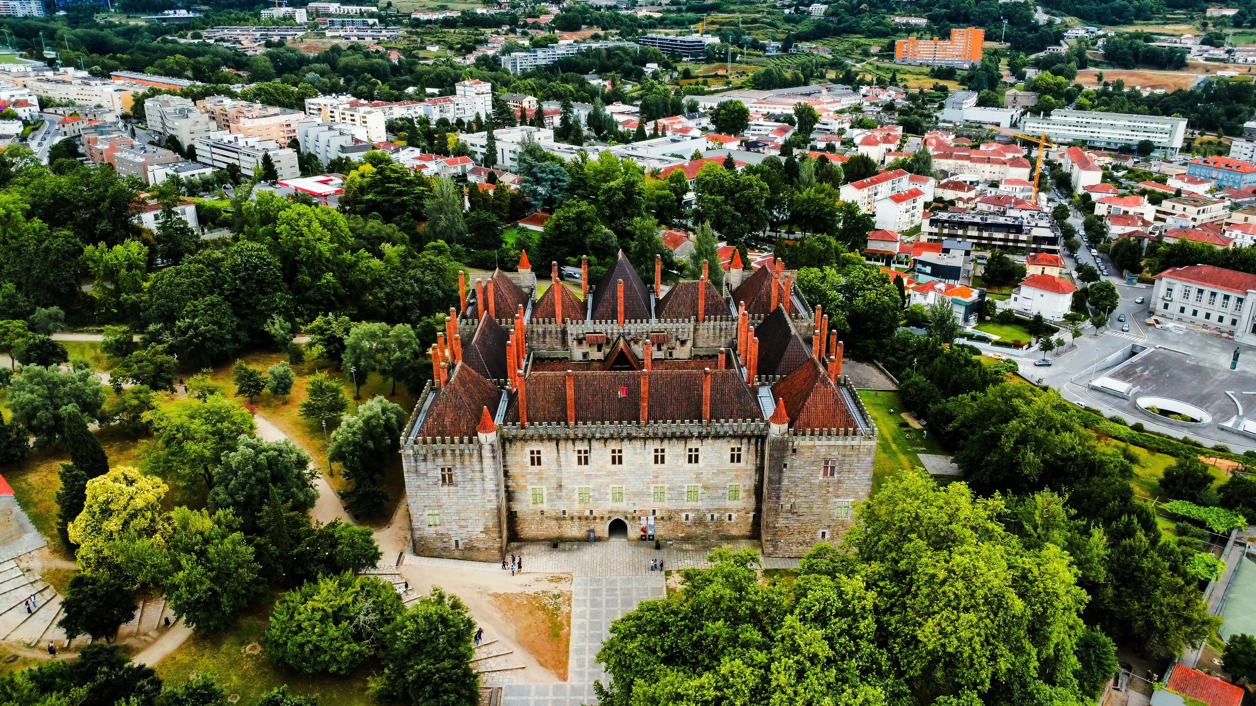 Aerial photograph of a turreted castle framed by dense greenery and a surrounding tree-lined town, captured from above to emphasize its scale and setting.