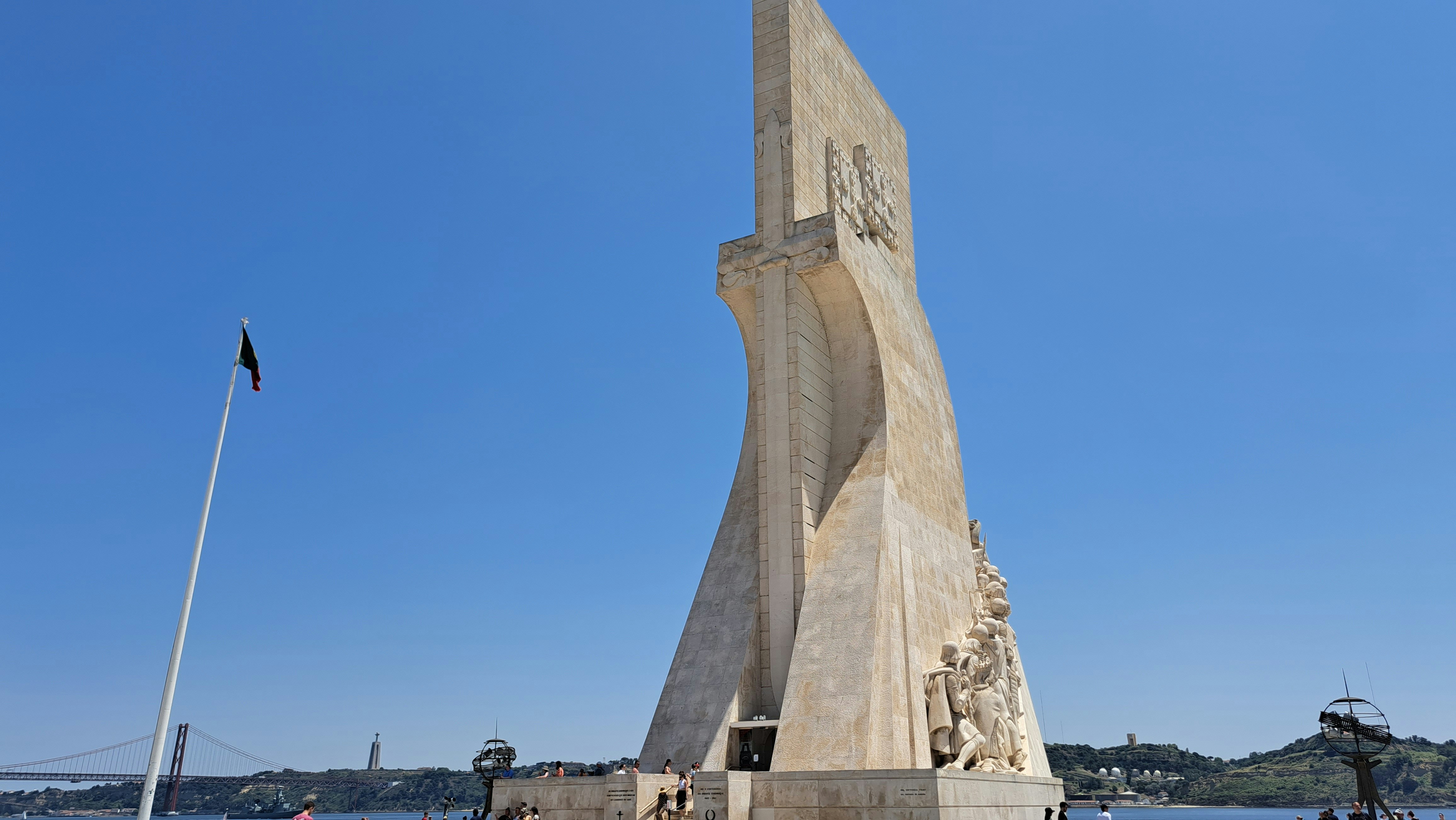 Tall stone monument with sculpted figures along its base rises against a clear blue sky. A flagpole stands to the left, emphasizing the waterfront setting.