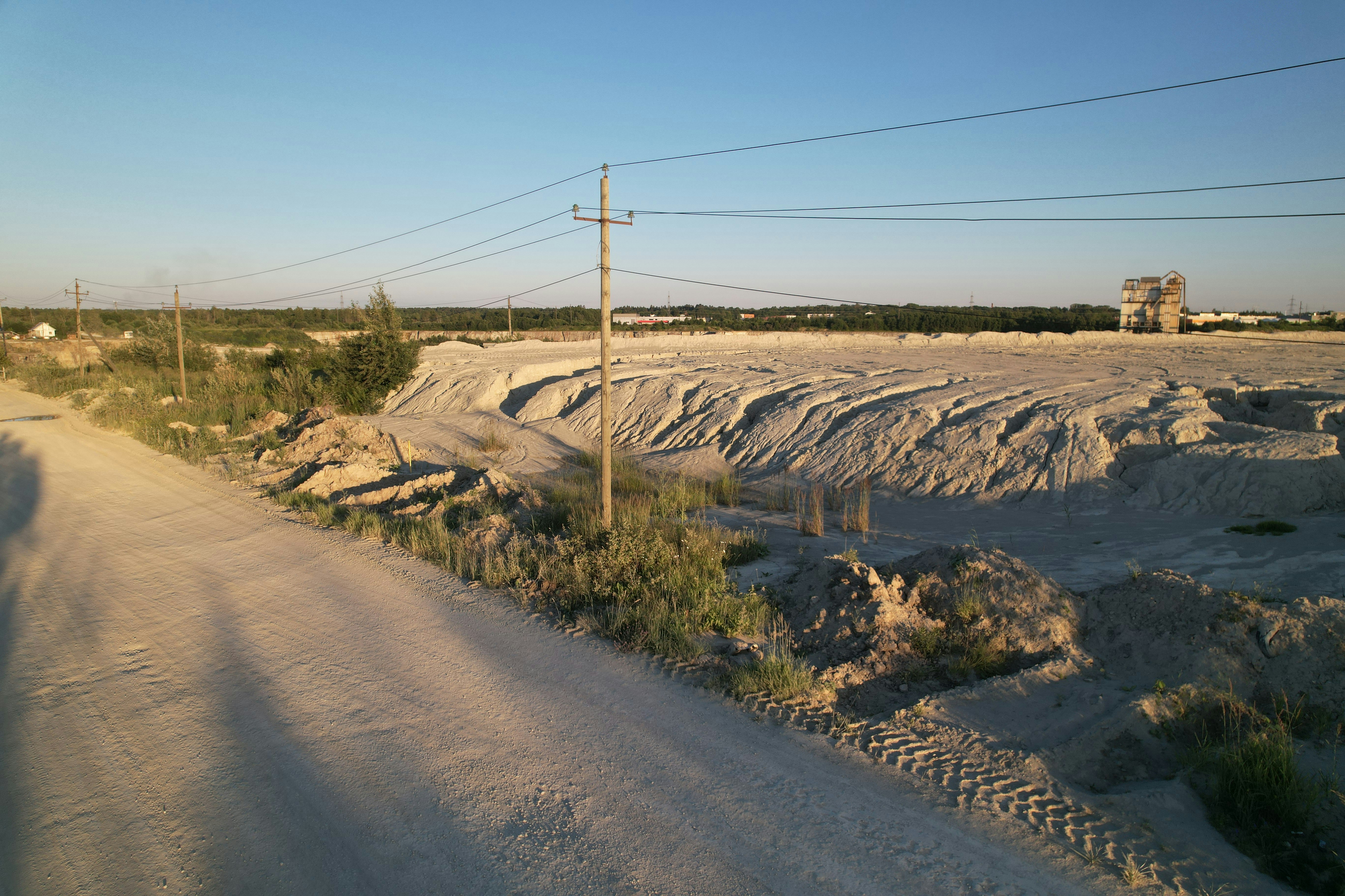 Dusty road leading alongside a sandy excavation site, with power lines stretching across the horizon. Industrial structures loom in the distance.